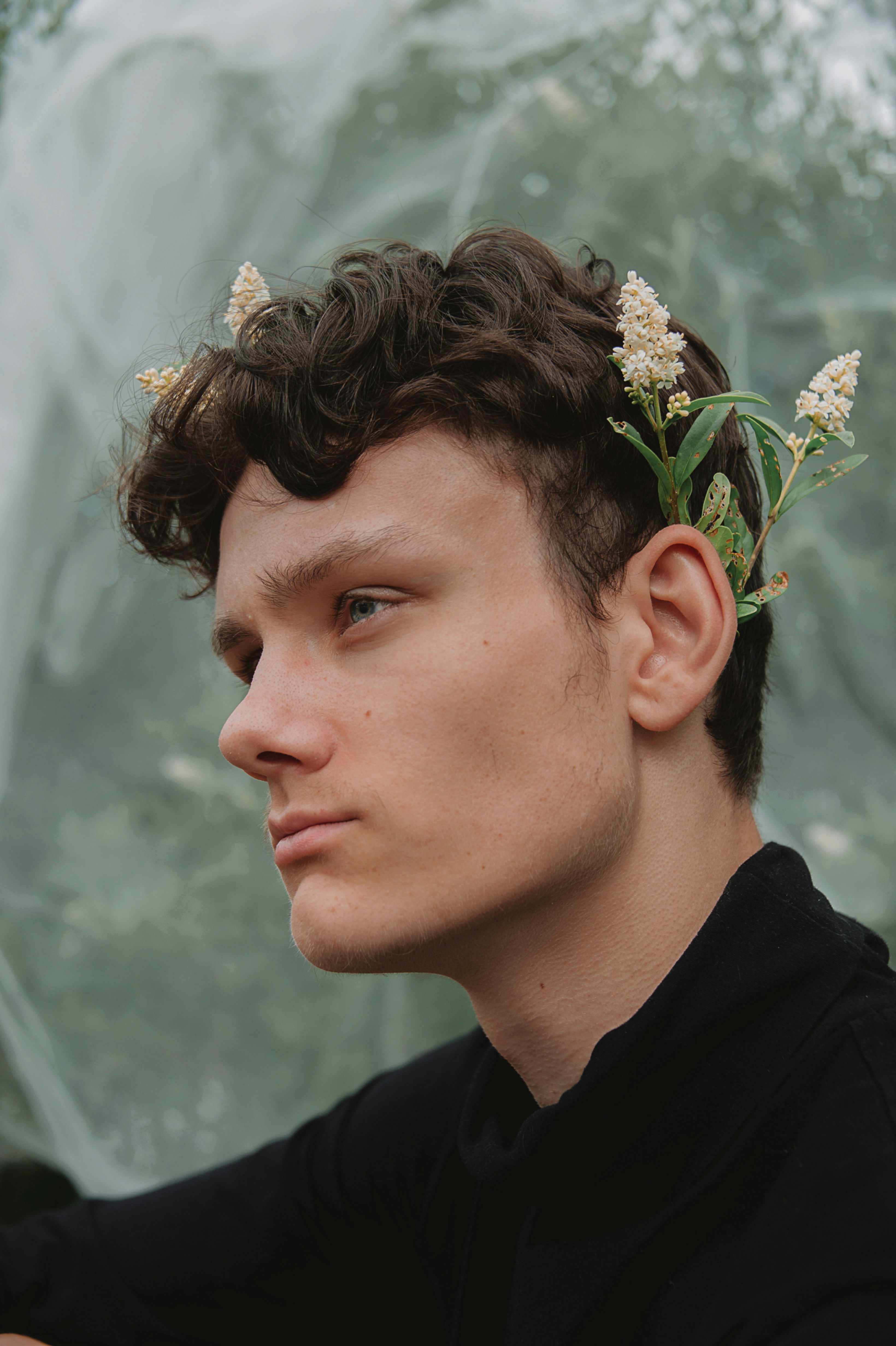 Profile photograph of a young man with a small floral crown, set against a soft greenhouse backdrop.