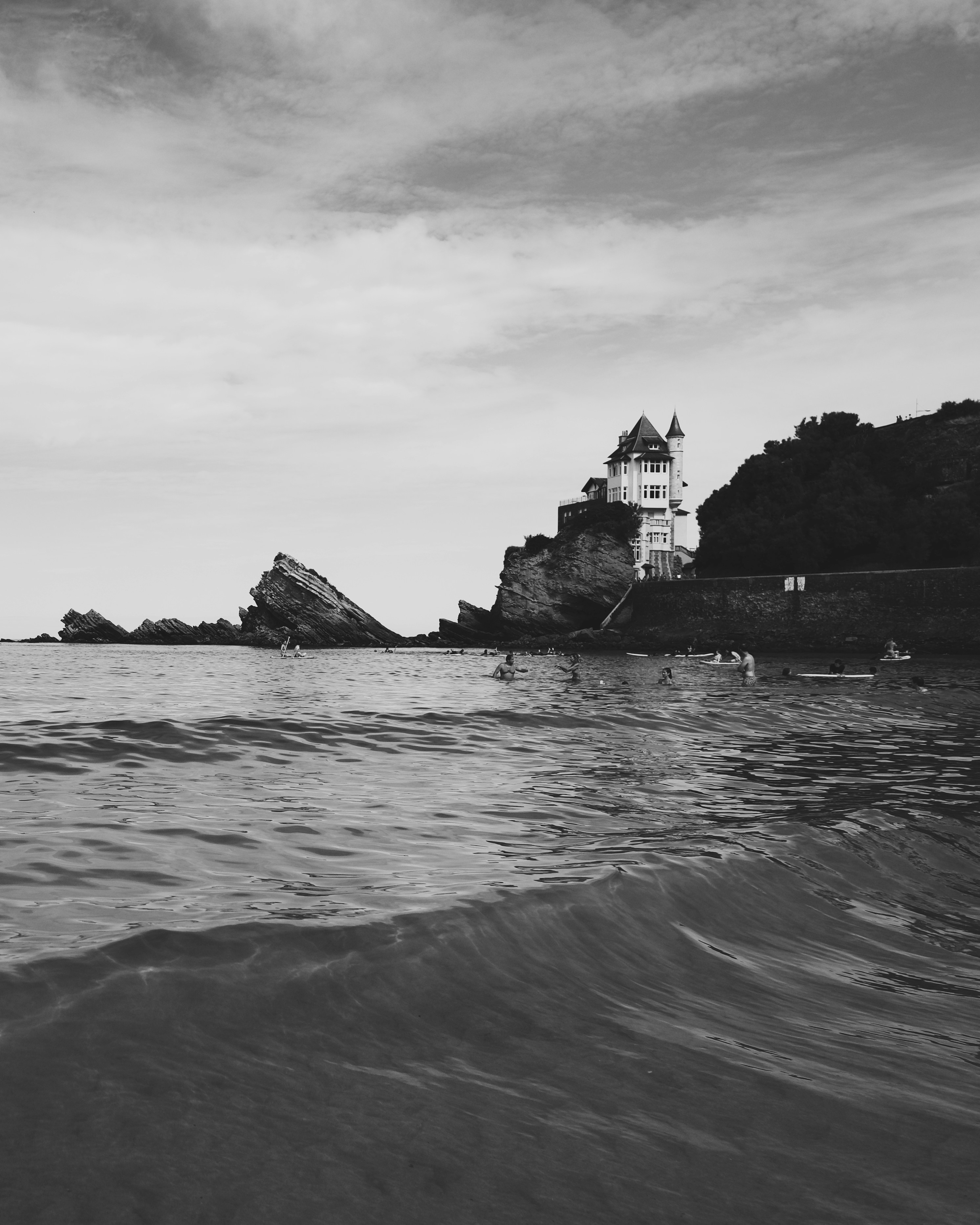 Historic coastal villa perched atop rocky cliffs, overlooking gentle waves and beachgoers. Black and white tones enhance the nostalgic atmosphere.