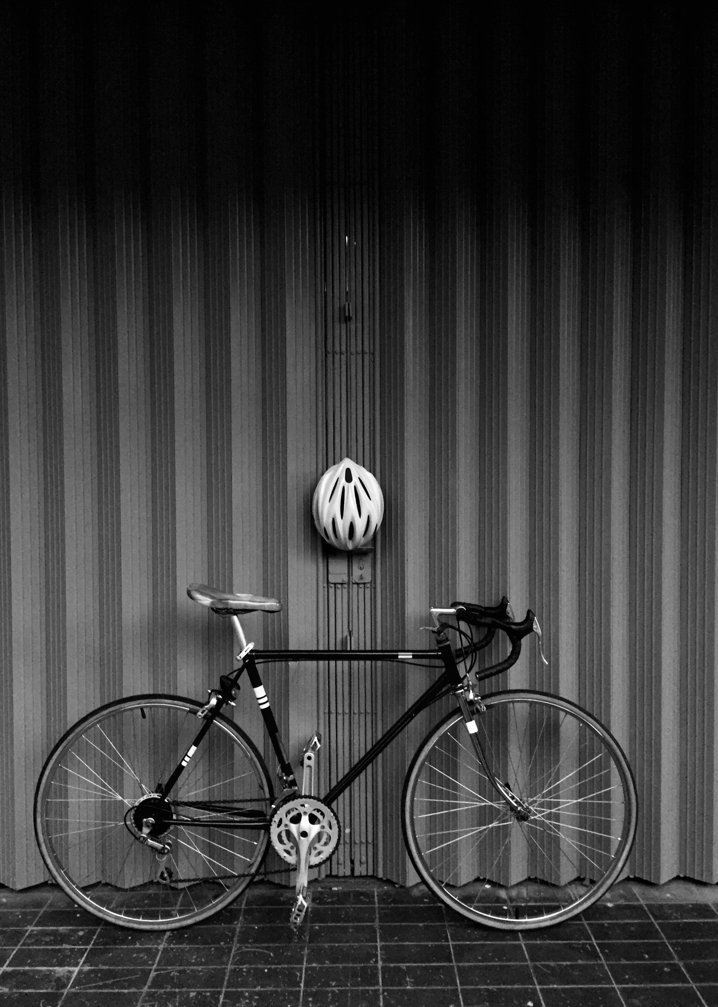 A black and white illustration of a bicycle leaning against a textured wall, with a helmet hanging above it. The simplicity emphasizes the relationship between the bike and its environment.