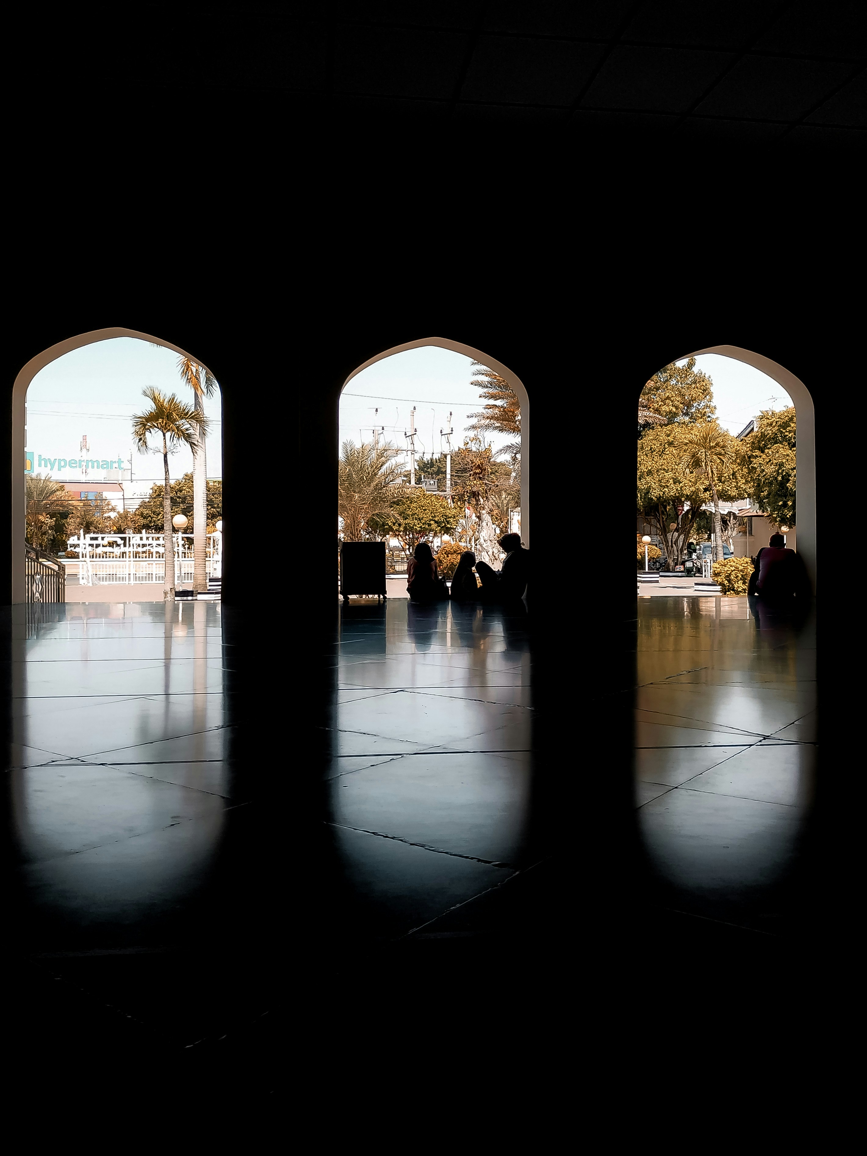 Silhouetted figures gather in a spacious corridor framed by three arches, revealing a vibrant outdoor scene beyond. The play of light and shadow creates a tranquil atmosphere.