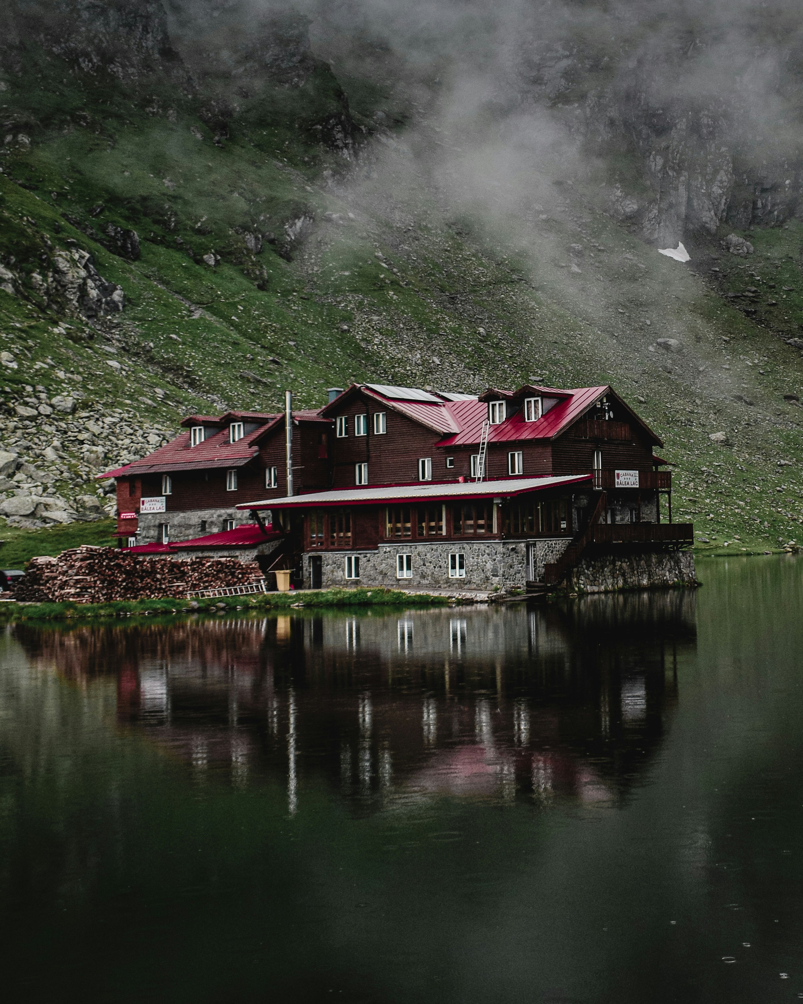 A rustic lodge nestled beside a tranquil lake, surrounded by misty mountains and lush greenery. The serene reflection in the water adds to the peaceful ambiance.