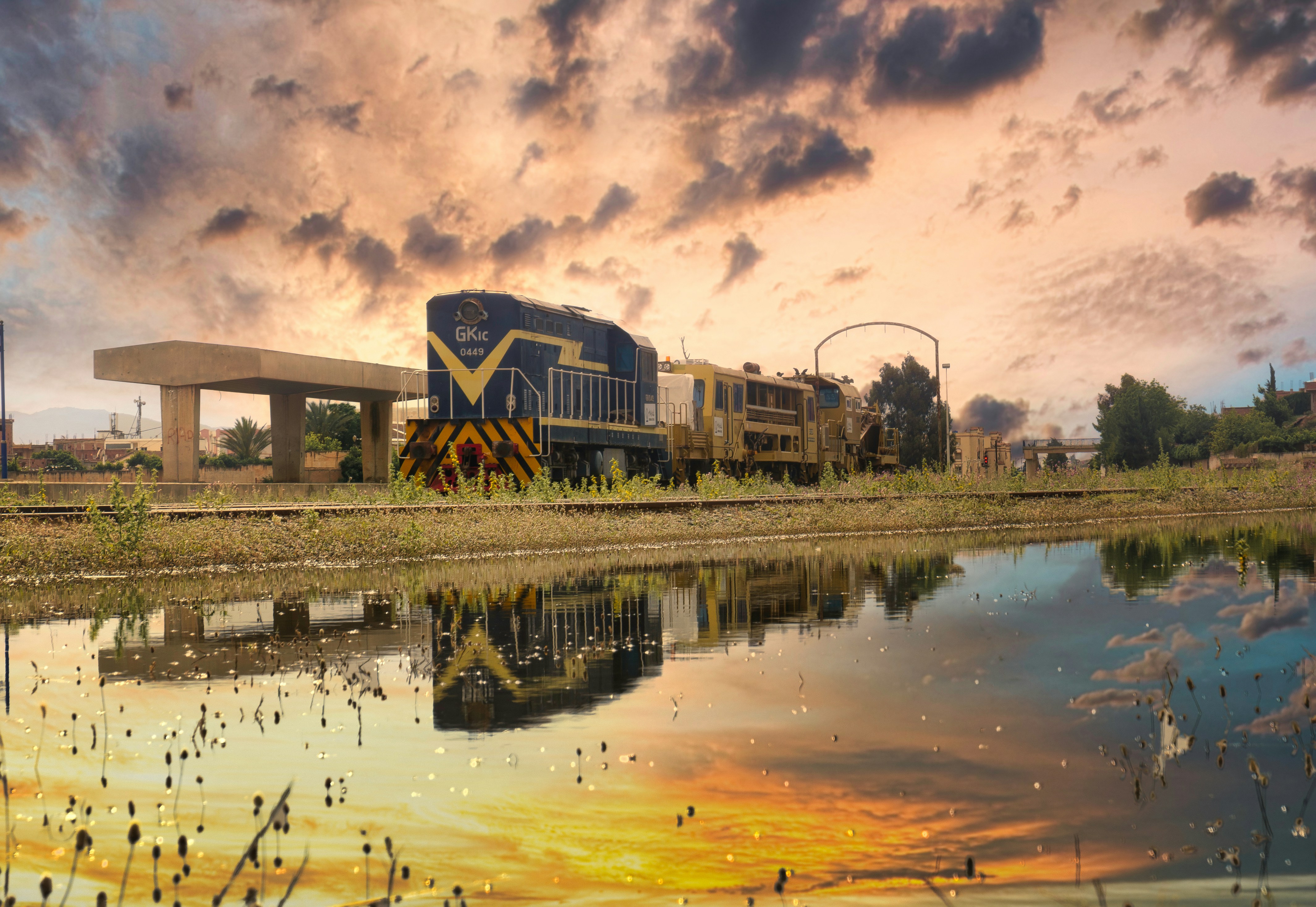 A vibrant train rests beside a reflective pool, framed by a dramatic sky at sunset. The scene captures the essence of industrial beauty and nature's tranquility.