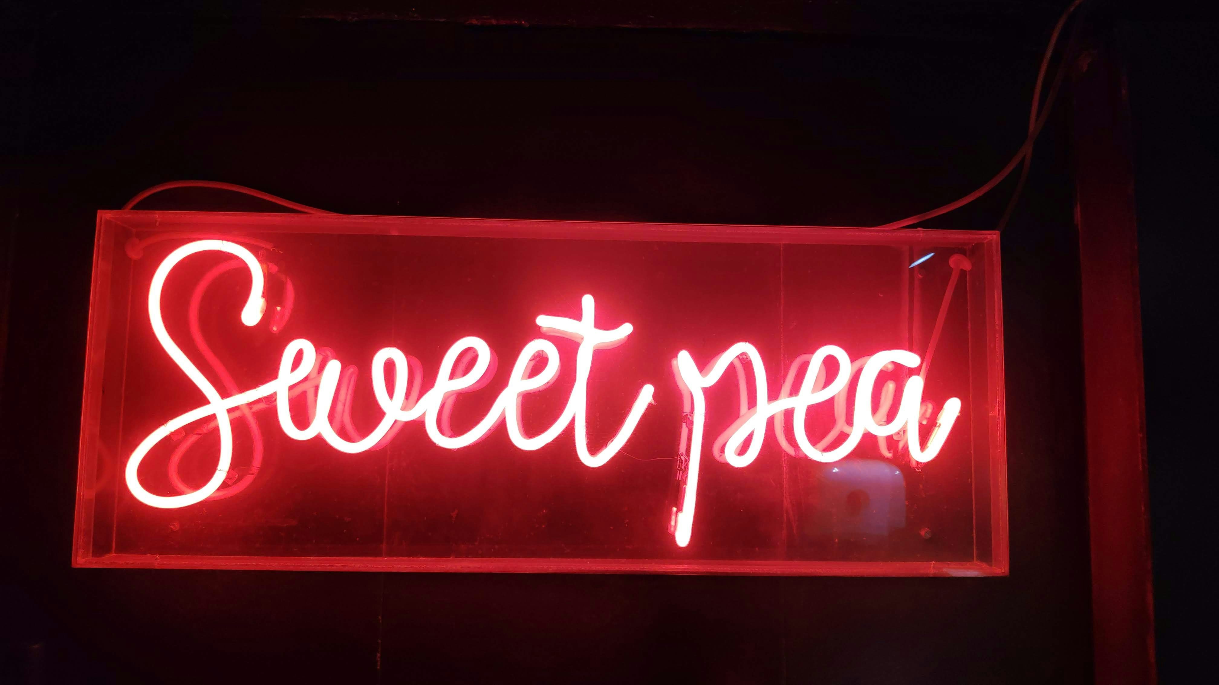 Red neon sign reading 'Sweet pea' in cursive, framed by a glowing rectangular panel against a dark wall.
