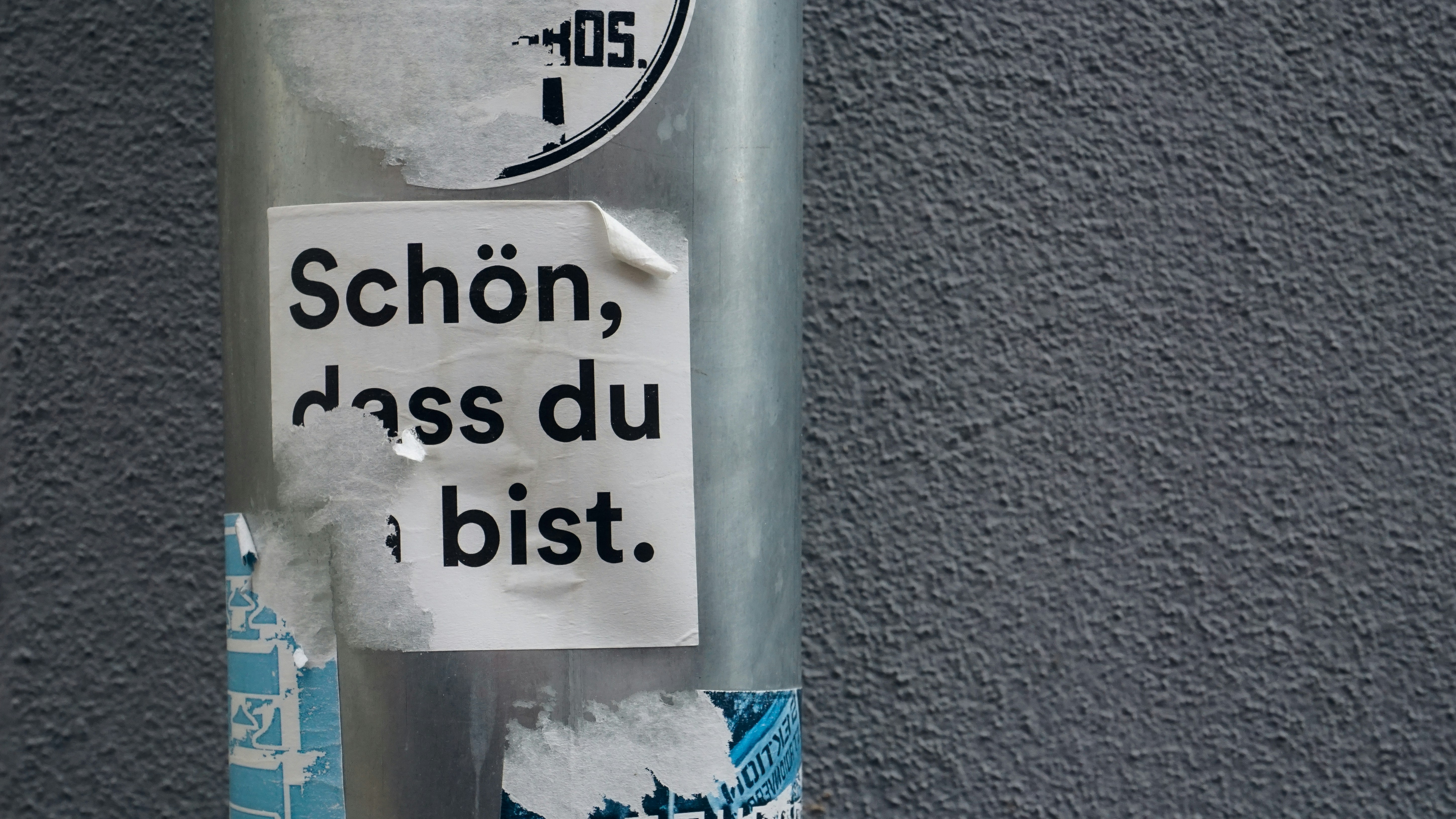 Sticker with German phrase on a weathered metal pole against a textured gray wall.