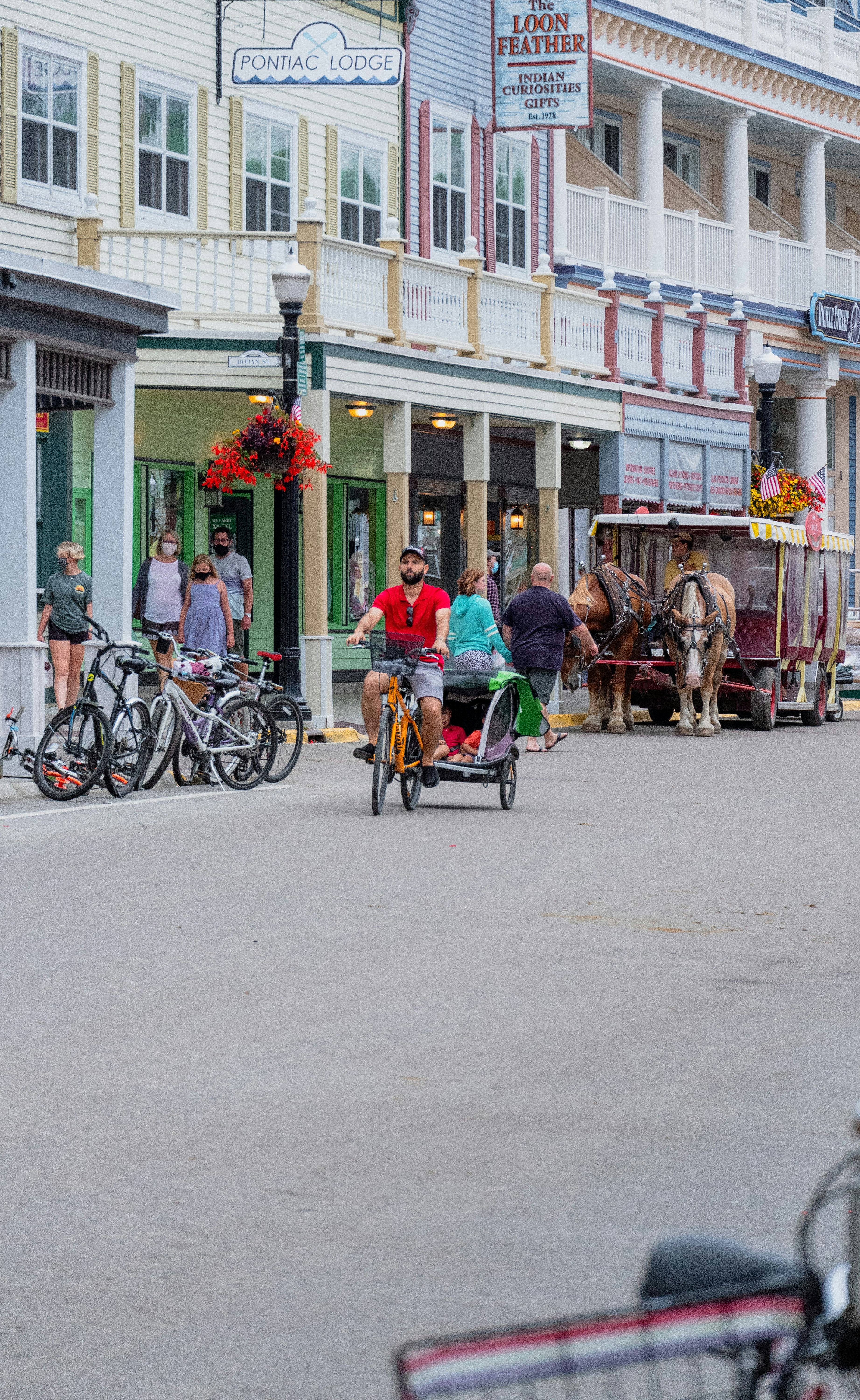 Busy street scene featuring cyclists, pedestrians, and a horse-drawn carriage amidst charming storefronts. Bright flowers add a touch of color to the lively atmosphere.