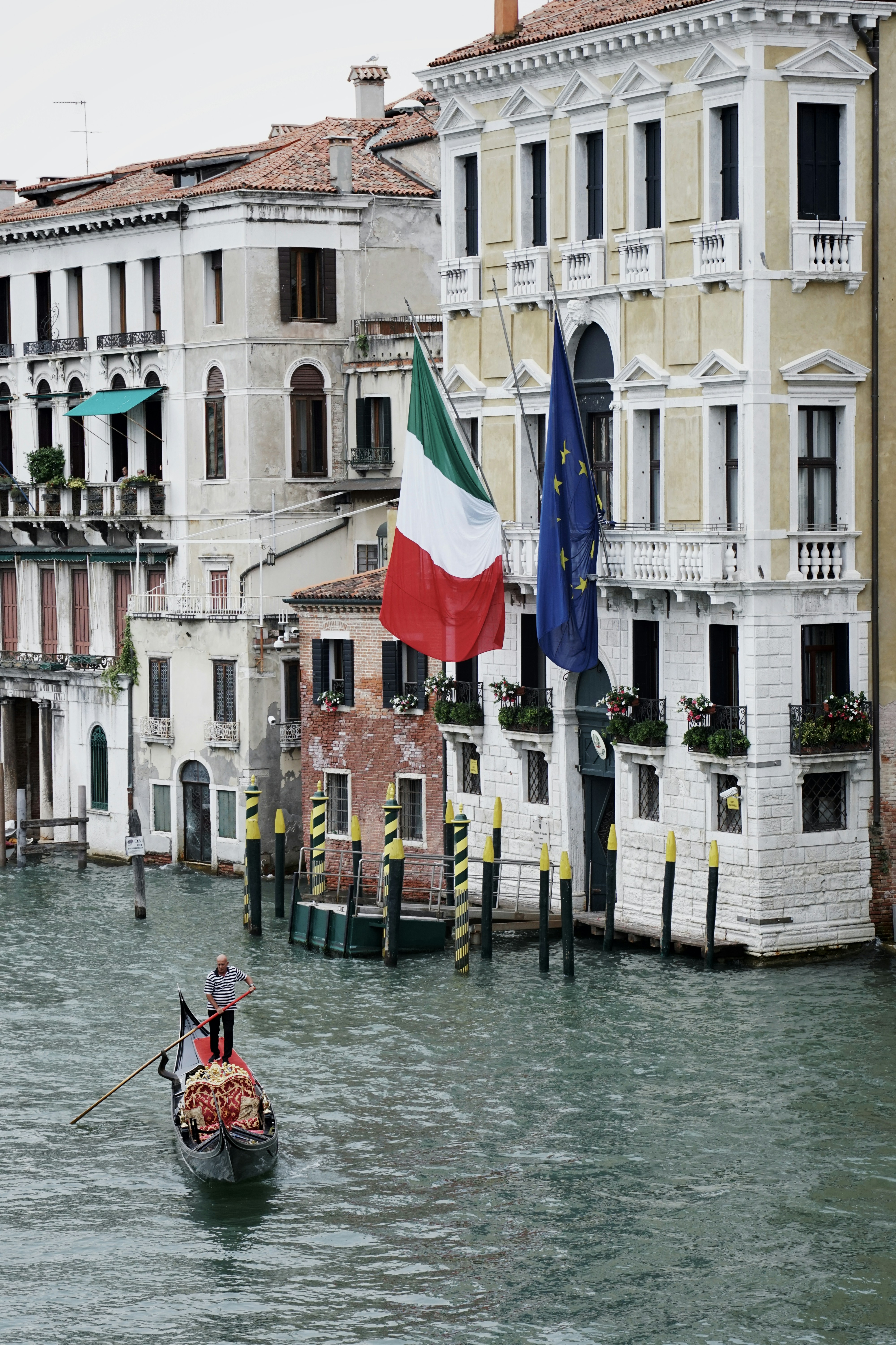 A gondolier navigates a canal in Venice, flanked by historic buildings and vibrant flags. The scene captures the essence of Venetian culture and life along the waterways.