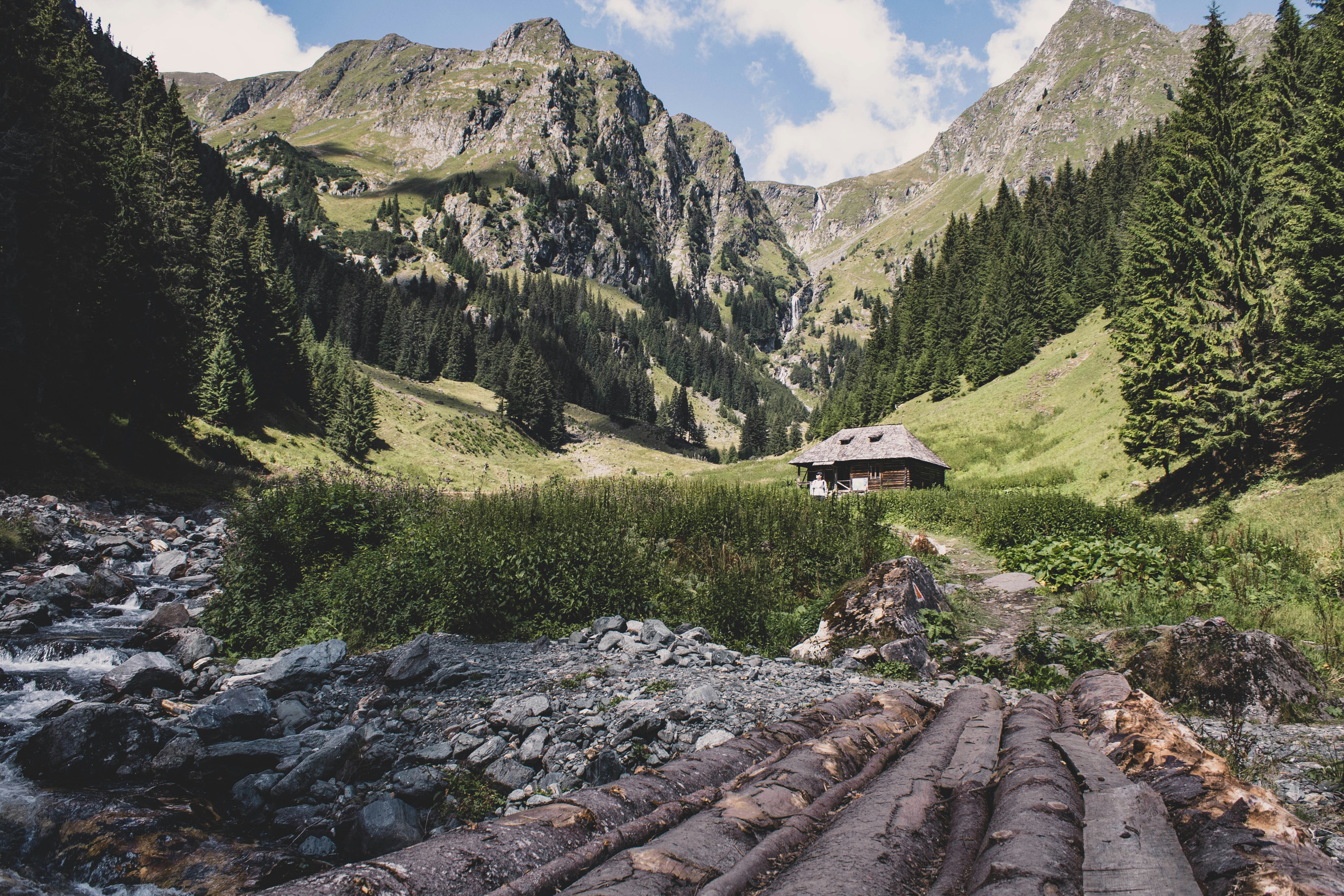 A rustic cabin nestled in a lush valley surrounded by towering mountains and dense forests, with a gentle stream flowing nearby.
