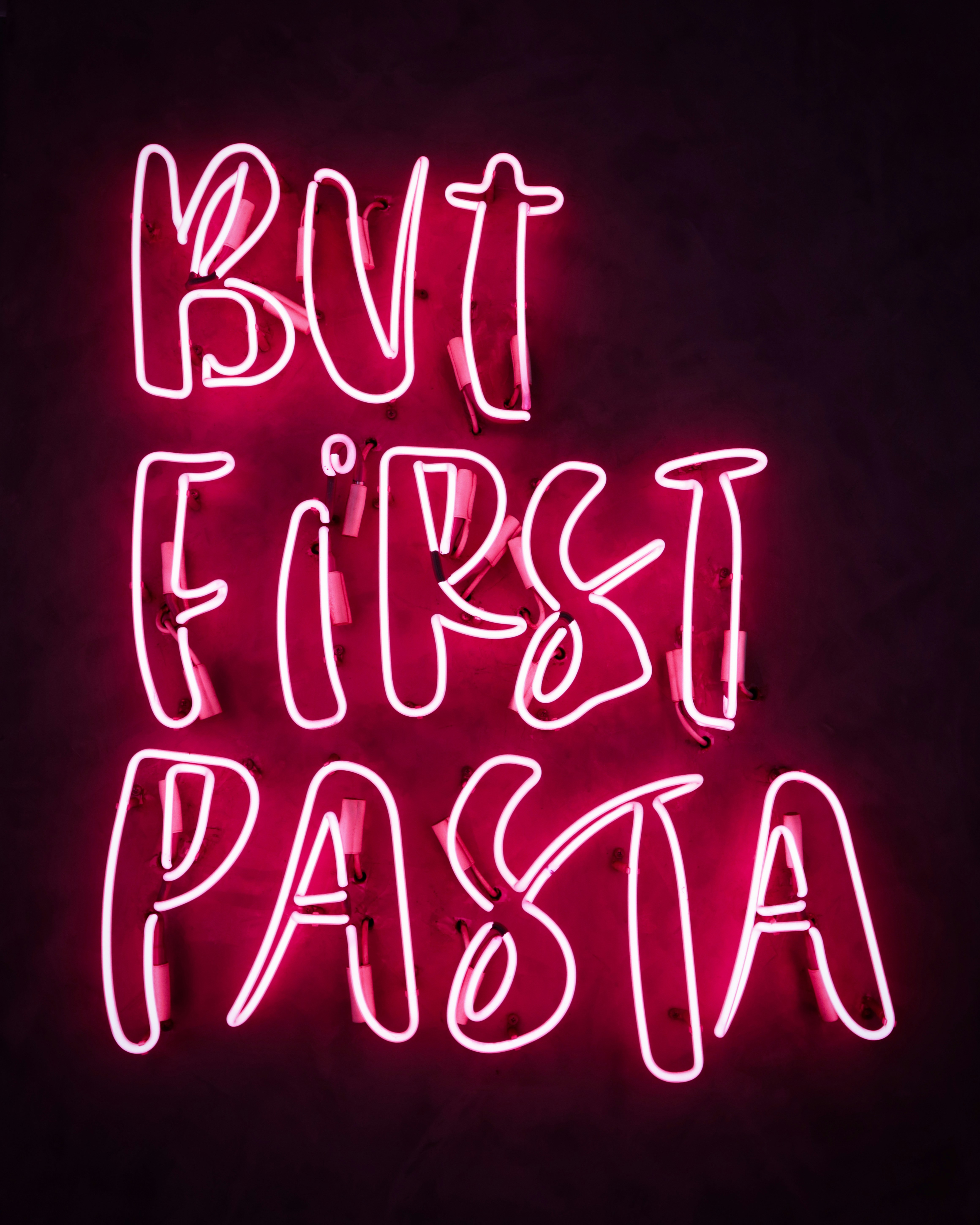 Pink neon sign reading 'BUT FIRST PASTA' against a dark background, radiating a bold glow and retro typography.