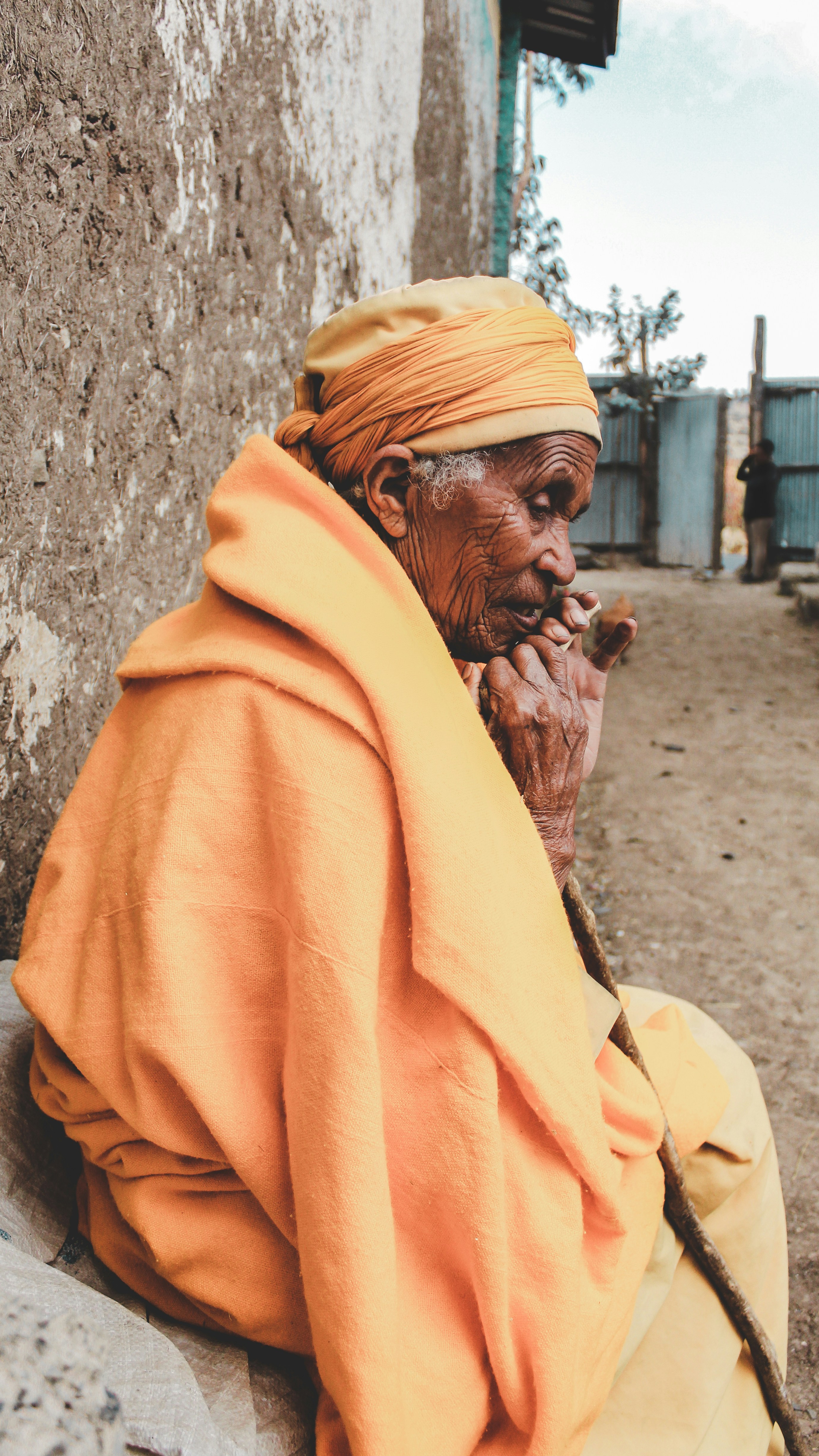 Elderly woman in vibrant orange attire seated against a rustic wall, holding a stick, lost in thought. The scene captures the essence of contemplation and heritage.