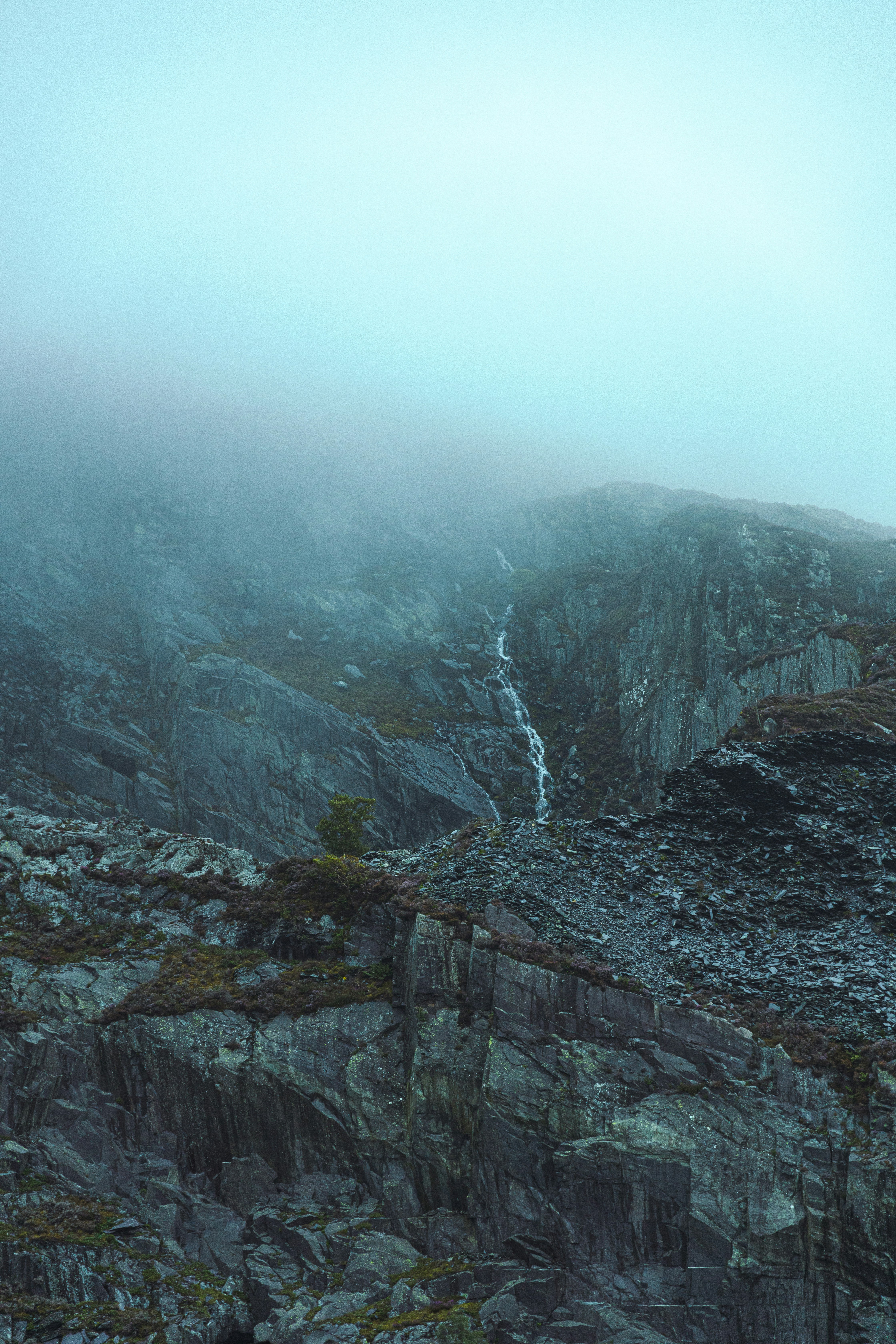 A serene mountain landscape shrouded in fog, featuring a subtle waterfall cascading down rocky terrain.
