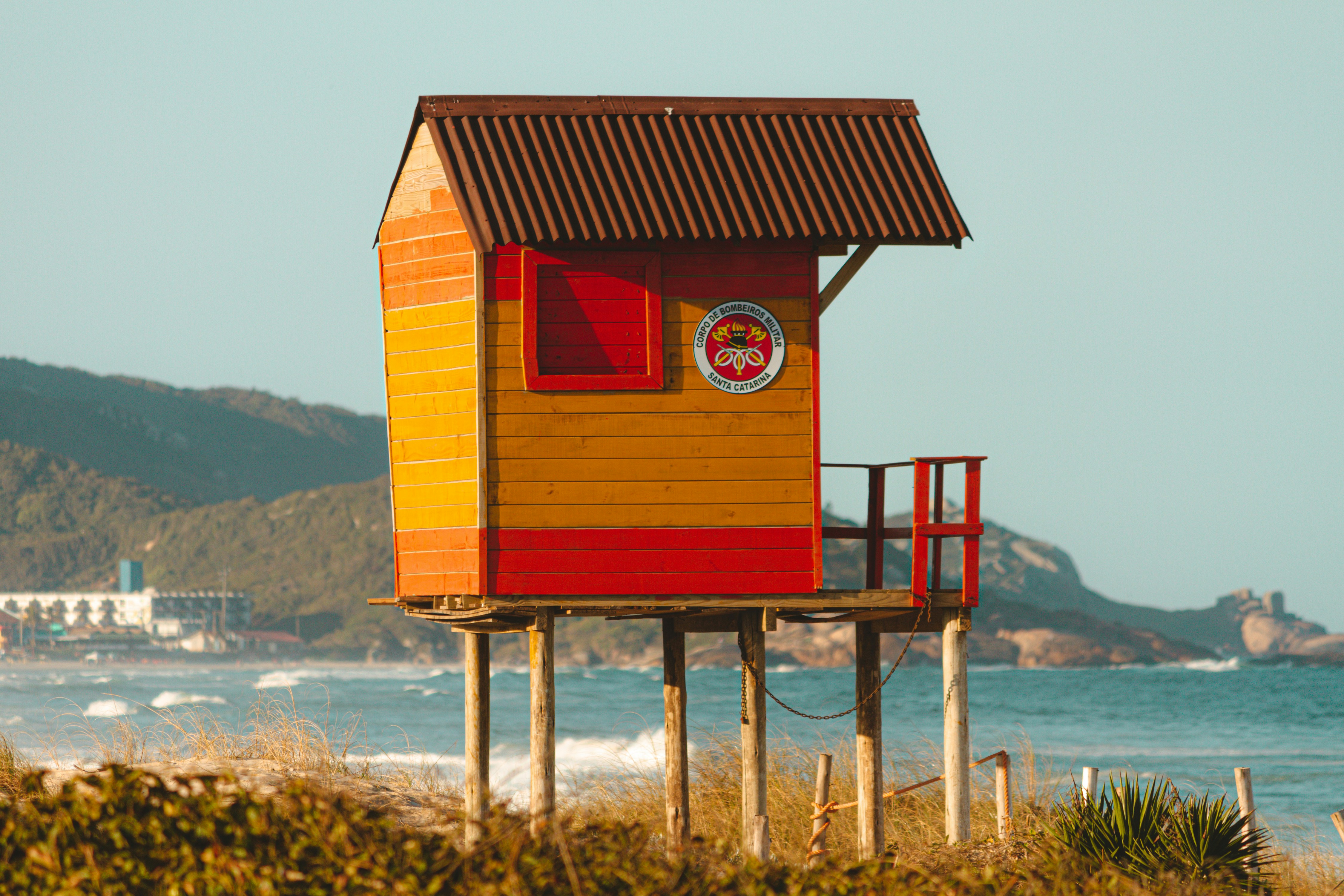 Colorful lifeguard hut perched on stilts by the beach, with rolling waves in the background. The structure features a prominent logo and bright hues.