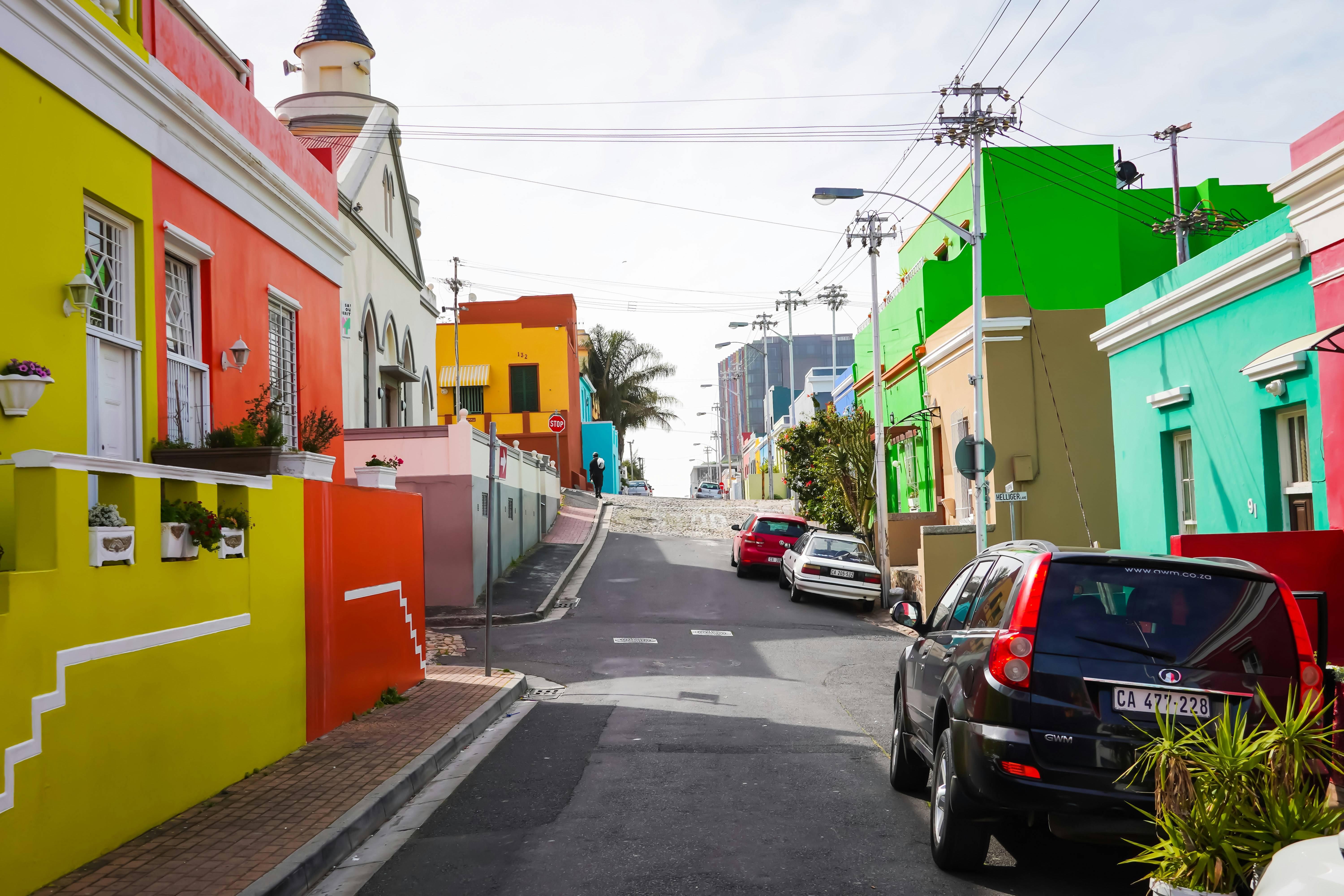 Brightly colored houses line a street in the historic Bo-Kaap neighborhood with parked cars and a clear blue sky overhead.