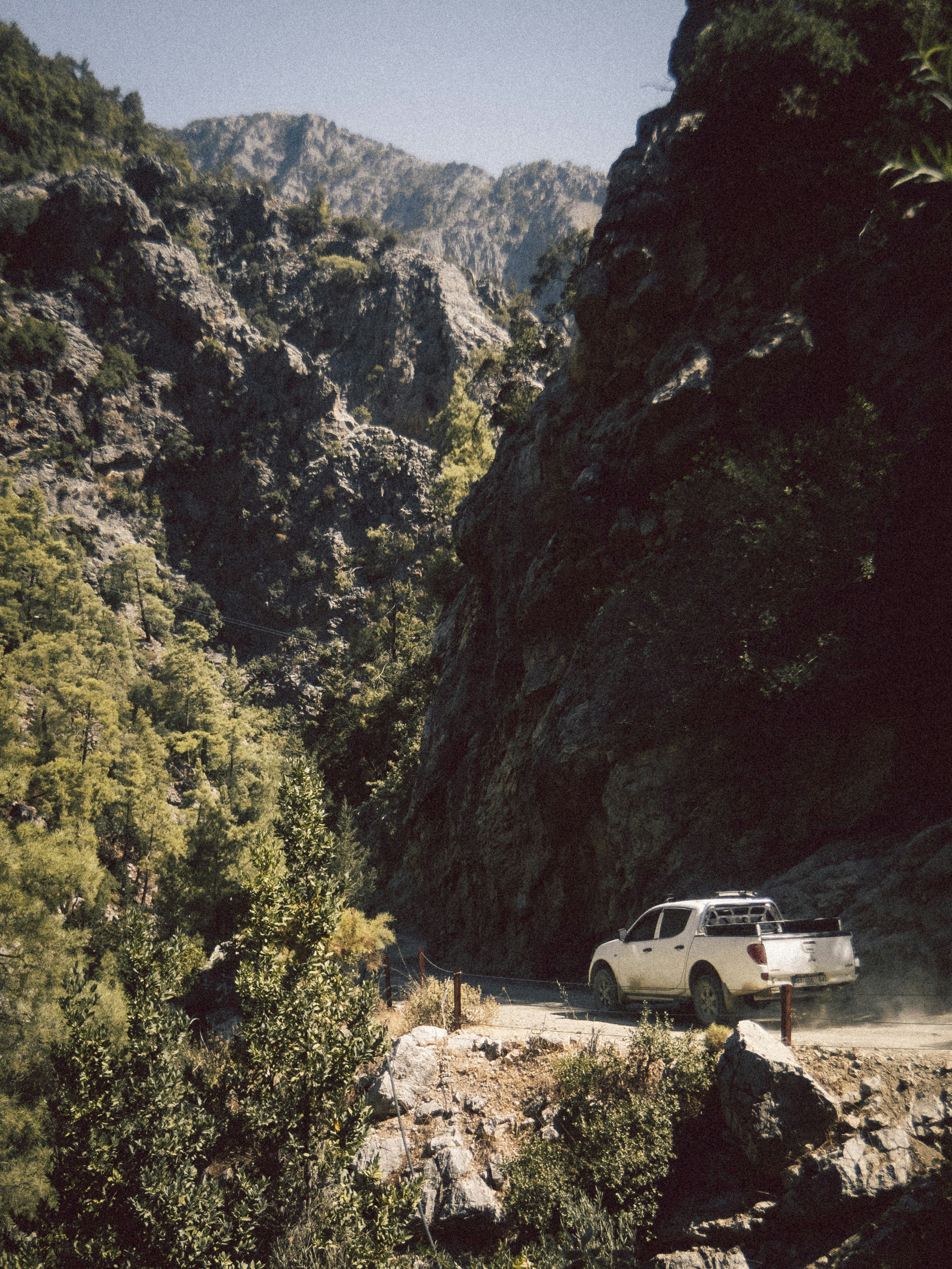 A white SUV navigates a winding dirt road flanked by towering rocky cliffs and lush greenery, highlighting the rugged beauty of the canyon landscape.