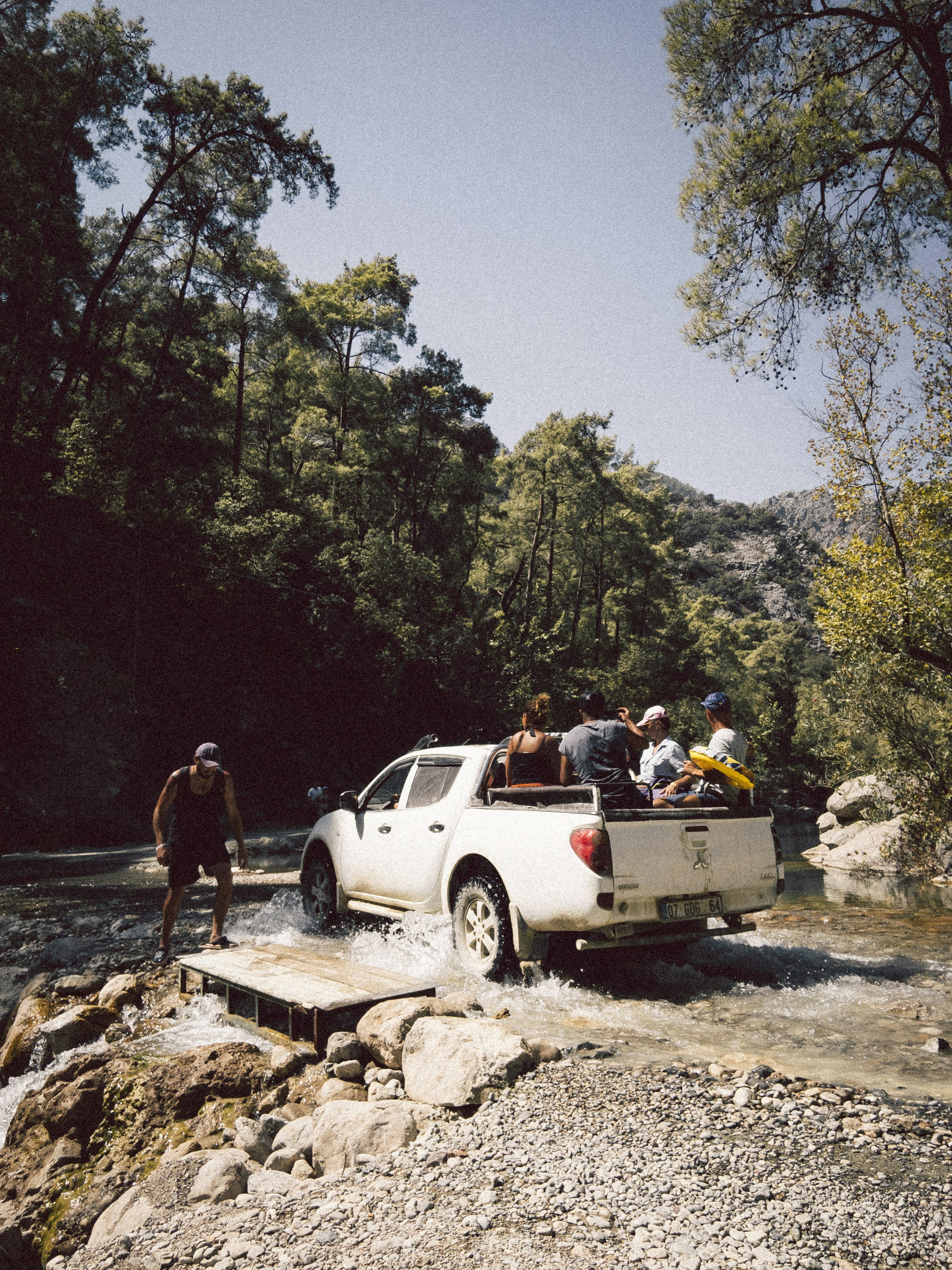 A white pickup truck navigates a shallow river with passengers enjoying the ride, surrounded by lush greenery and rocky terrain.