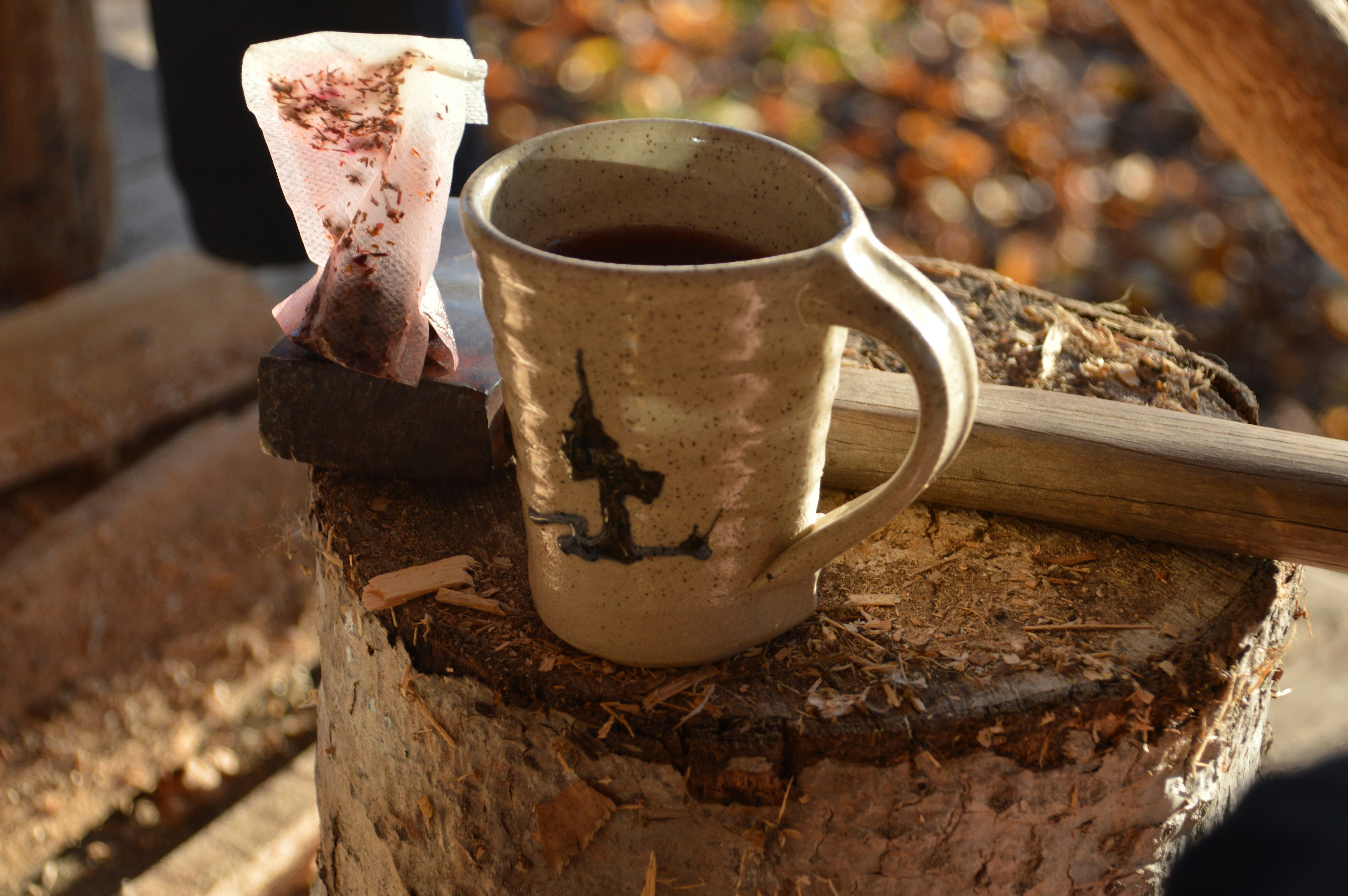 Ceramic mug filled with tea rests on a wooden stump beside a used tea bag and an axe, capturing a tranquil outdoor setting.
