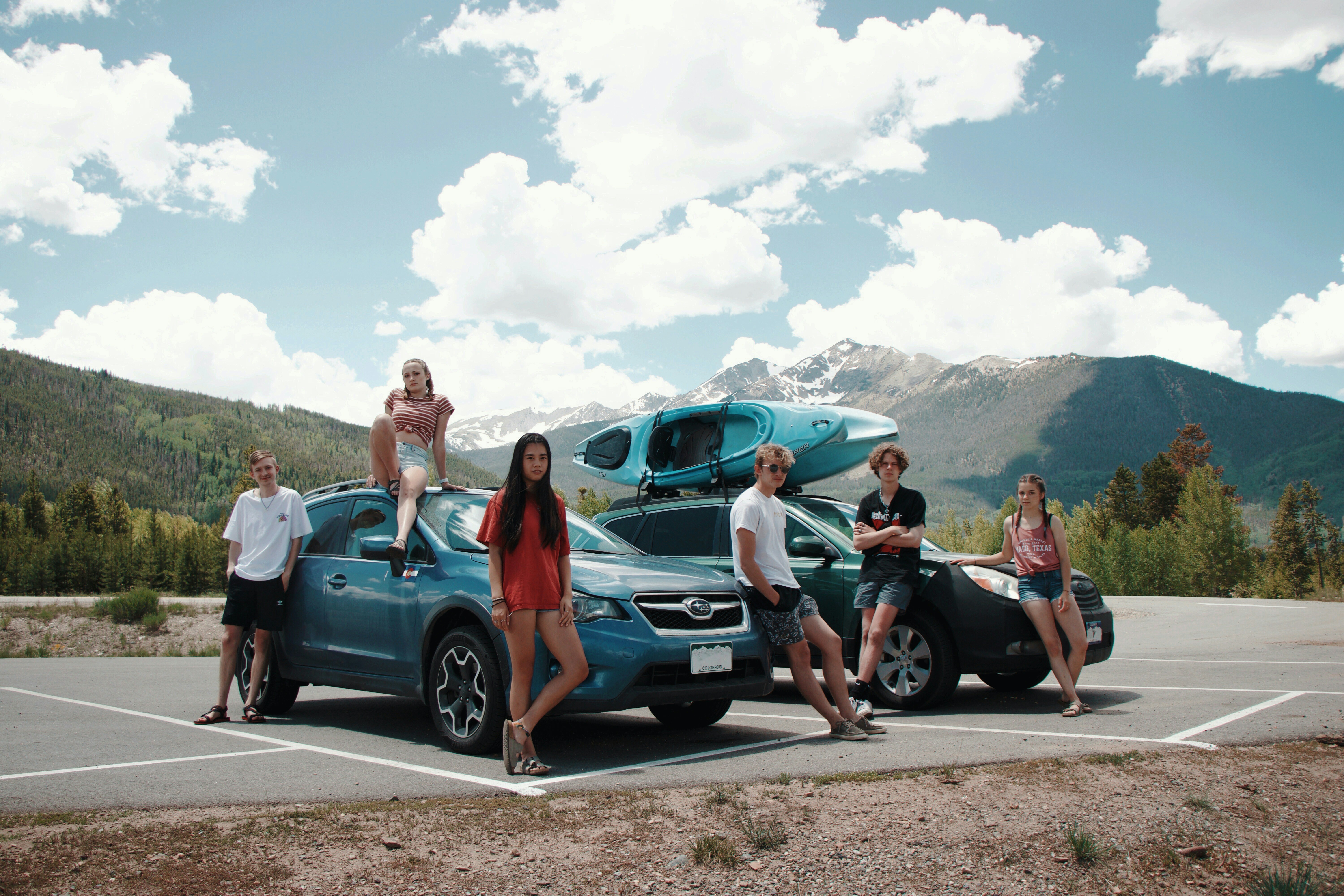 Group of friends posing with two cars in a mountainous parking area, with kayaks on top and a backdrop of lush greenery and clouds.