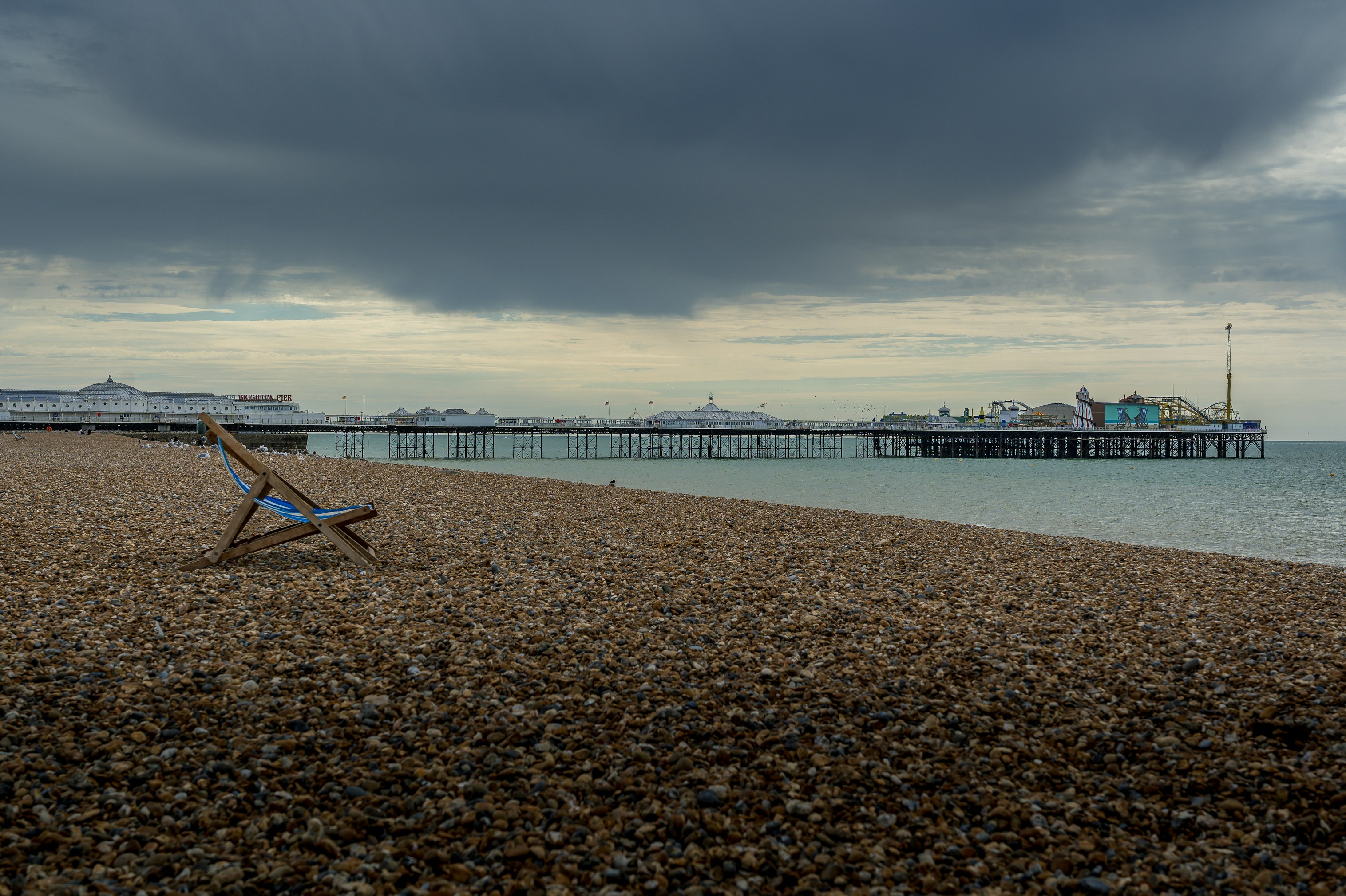 Pebble Beach Recline: A Lone Deck Chair Framed by a Distant Pier Under a Moody Sky