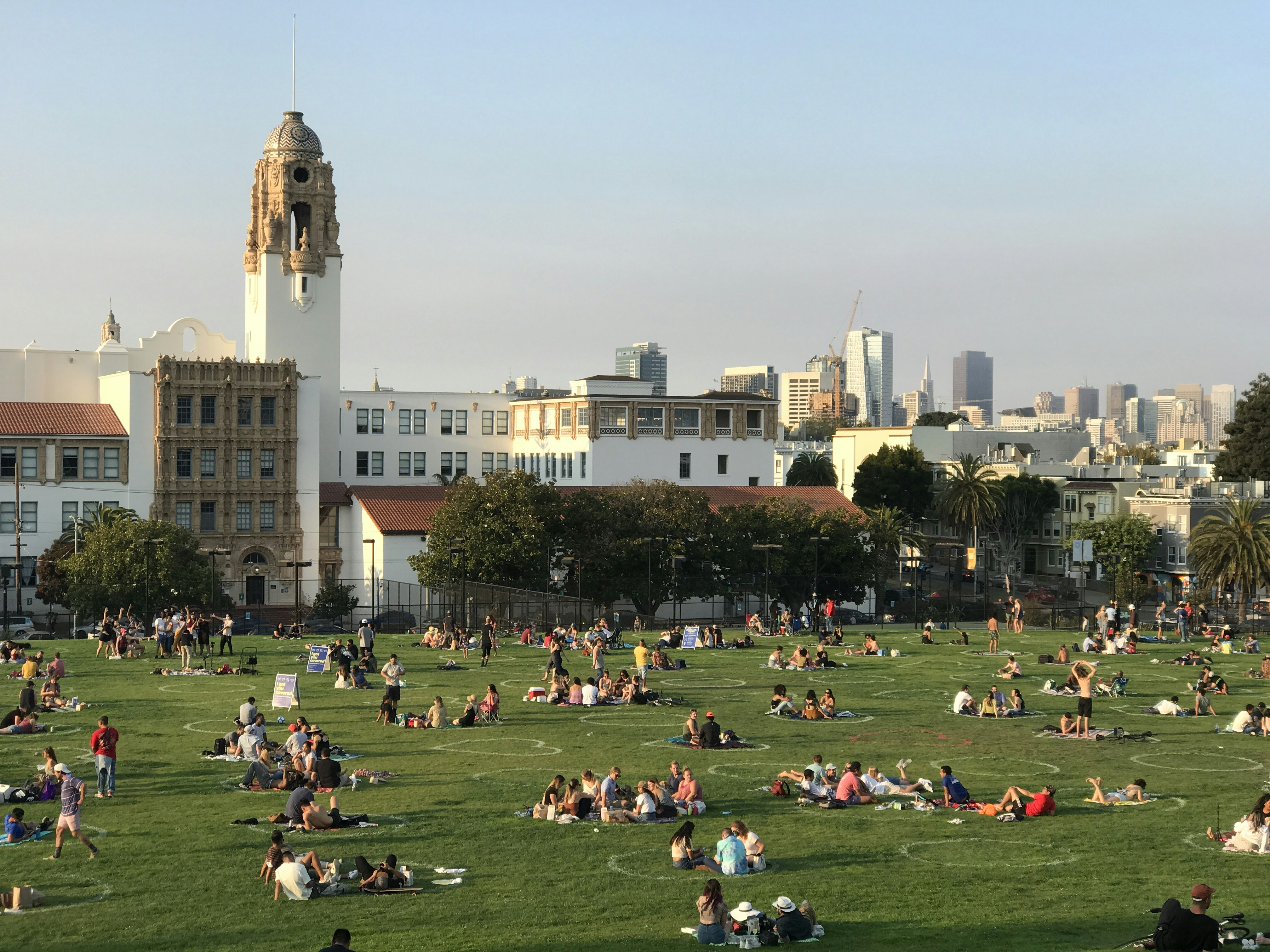 People relaxing on a grassy field near a historic tower with a city skyline in the background.