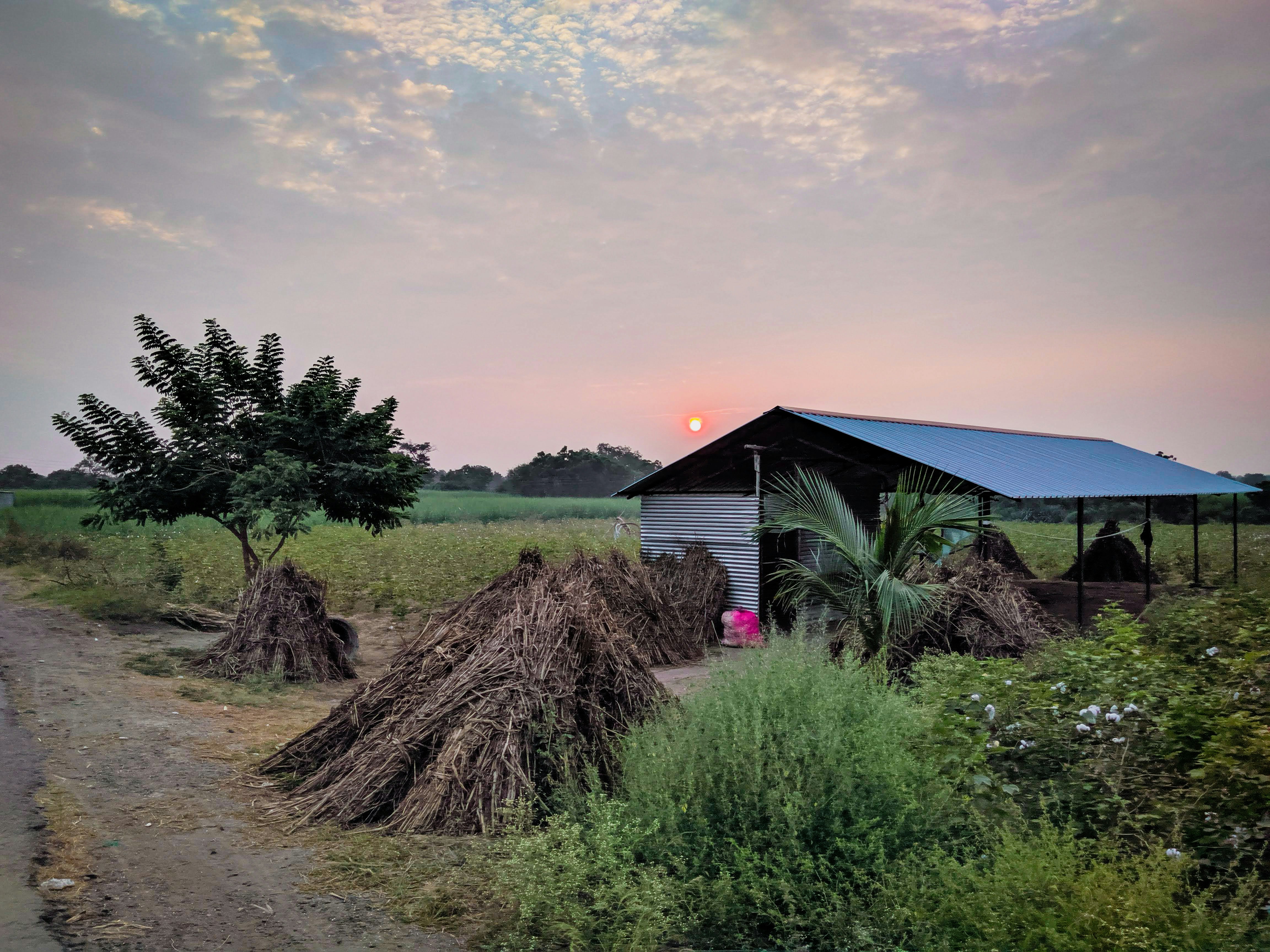 Rural farm scene at sunset with a small shed, stacked hay, and a dirt path winding through green fields beneath a pastel sky.