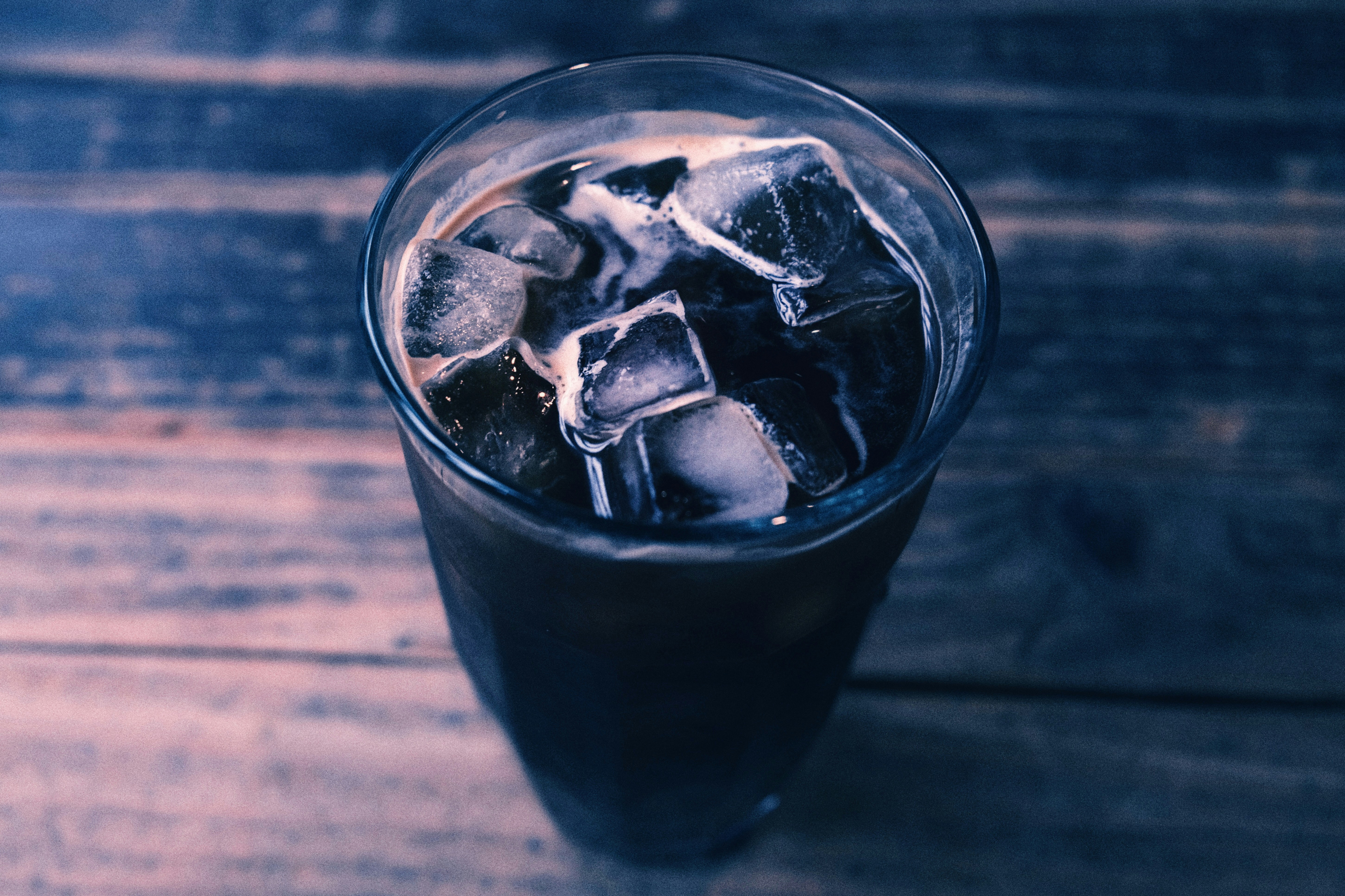 A tall glass filled with dark beverage and ice cubes resting on a rustic wooden surface.