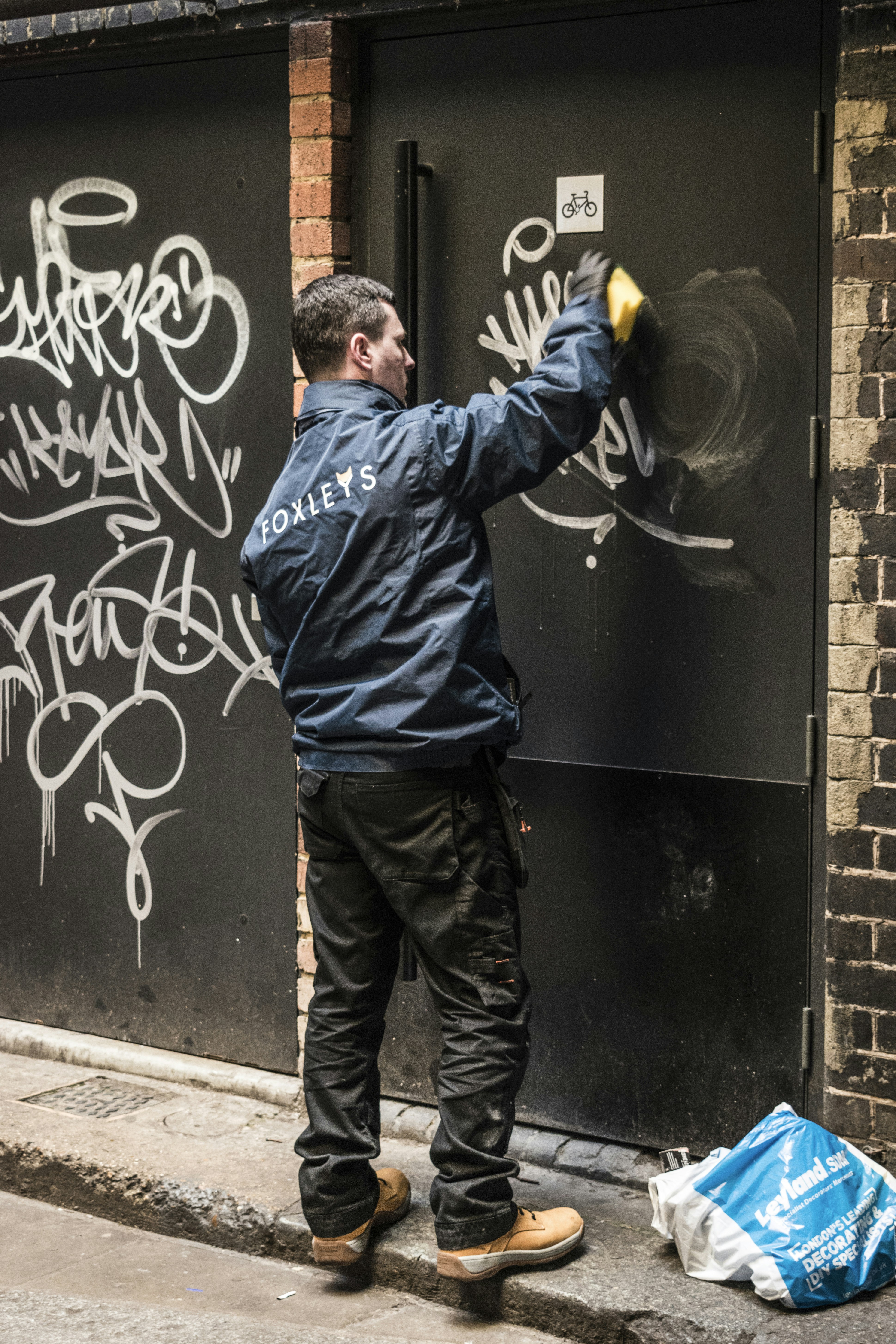A man in a blue jacket removes graffiti from a black door in an alleyway, showcasing the contrast between art and maintenance.