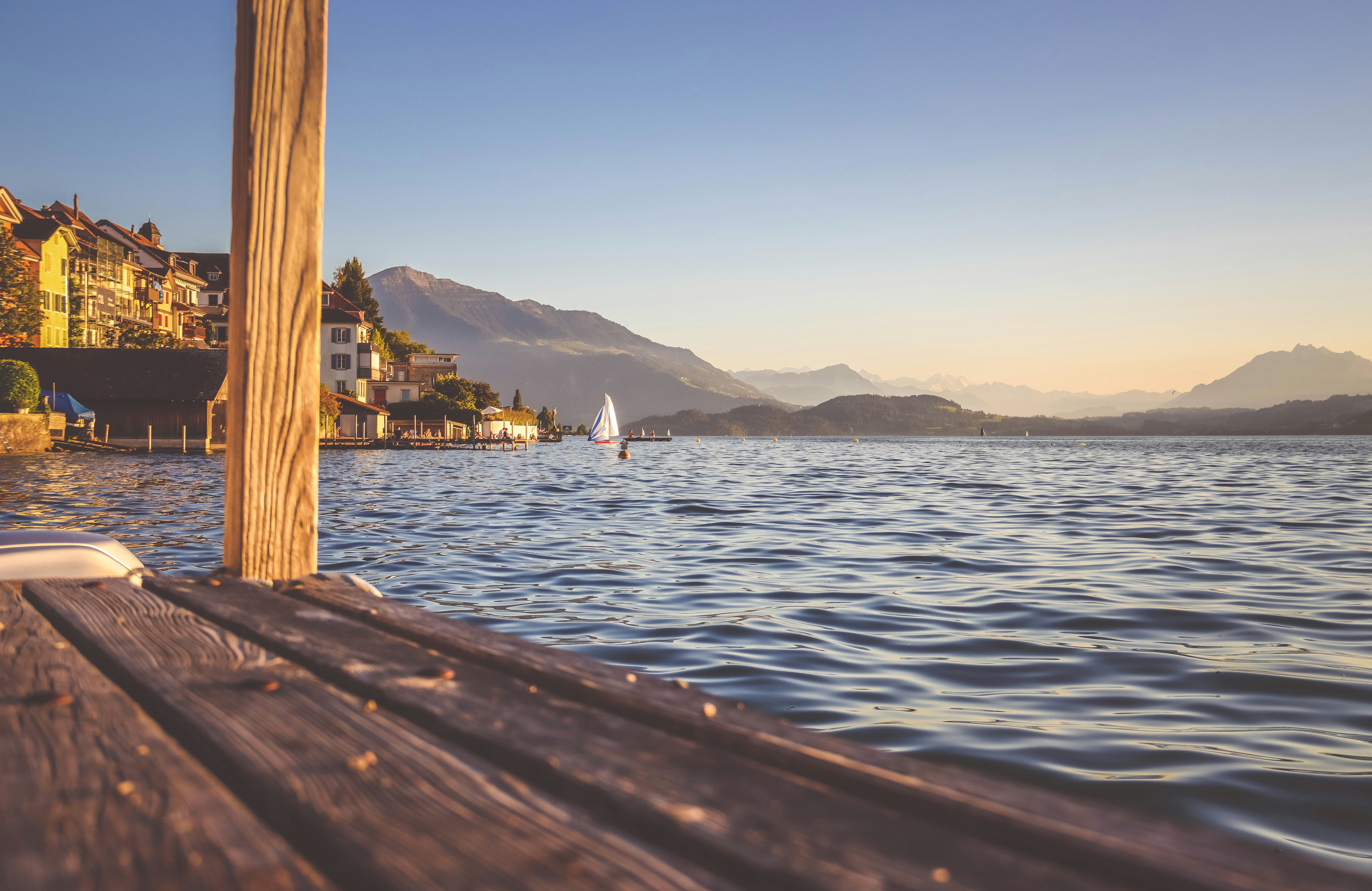 Calm lake scene with a wooden pier in the foreground and a sailboat gliding across the water under a golden sunset.