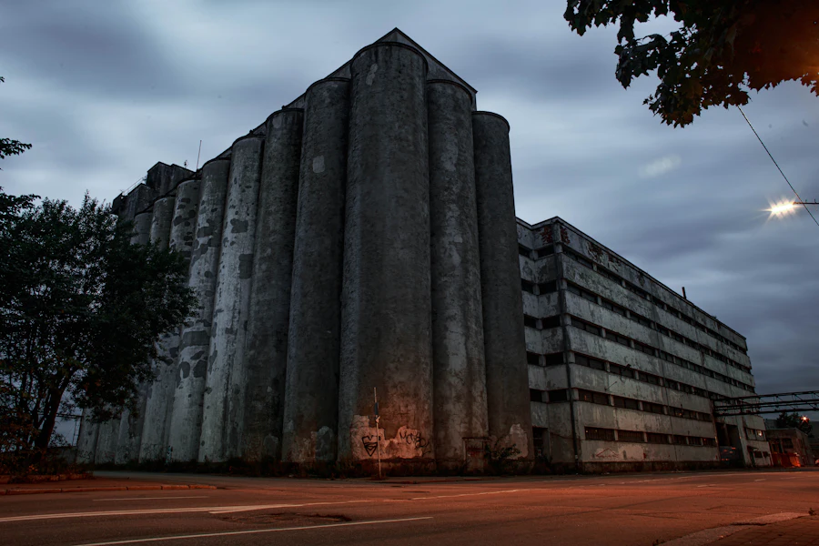 Weathered grain silos dominate a dim street corner at dusk; the scene is gritty with a moody blue hour tone and subtle red street lighting highlighting textures. Industrial building with concrete towers stands as the centerpiece.