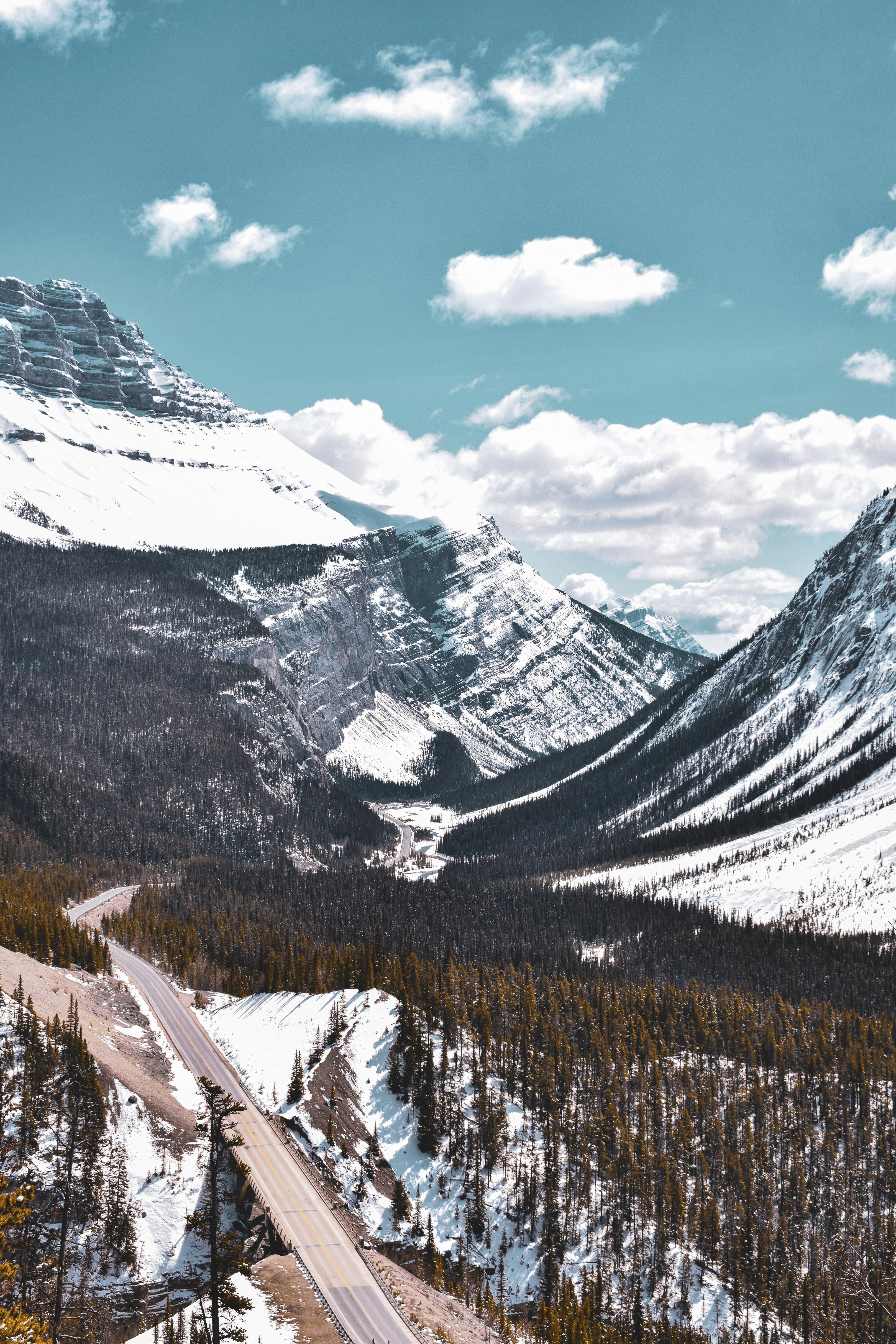 Snow-covered mountain peaks loom over a winding road cutting through a lush forest valley, framed by dramatic skies. 