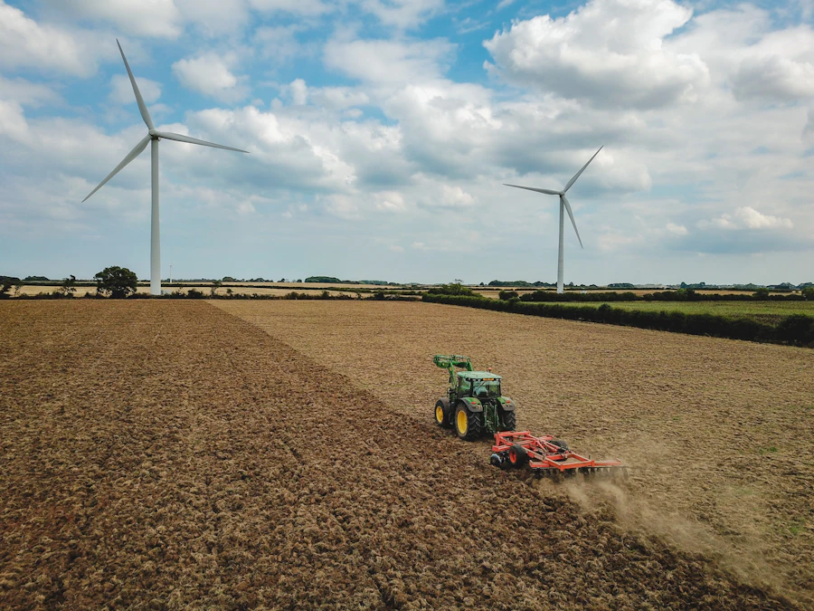 Dusty tractor work moves through a plowed field beneath tall wind turbines, captured from above to emphasize scale and rural rhythm. This is a drone shot with a broad landscape layout.