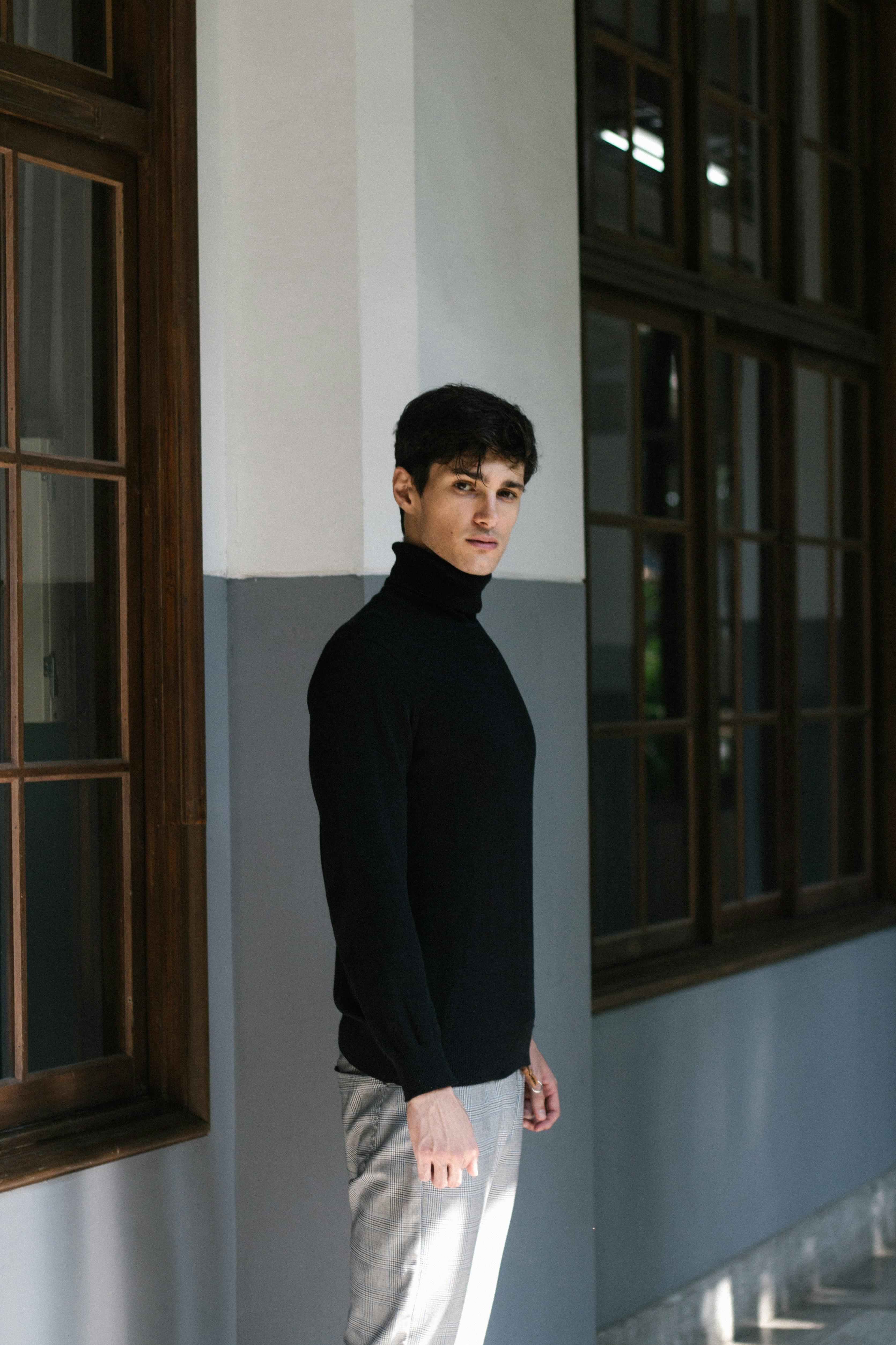 Young man in a black turtleneck stands against a backdrop of large windows, exuding a sense of calm and introspection.