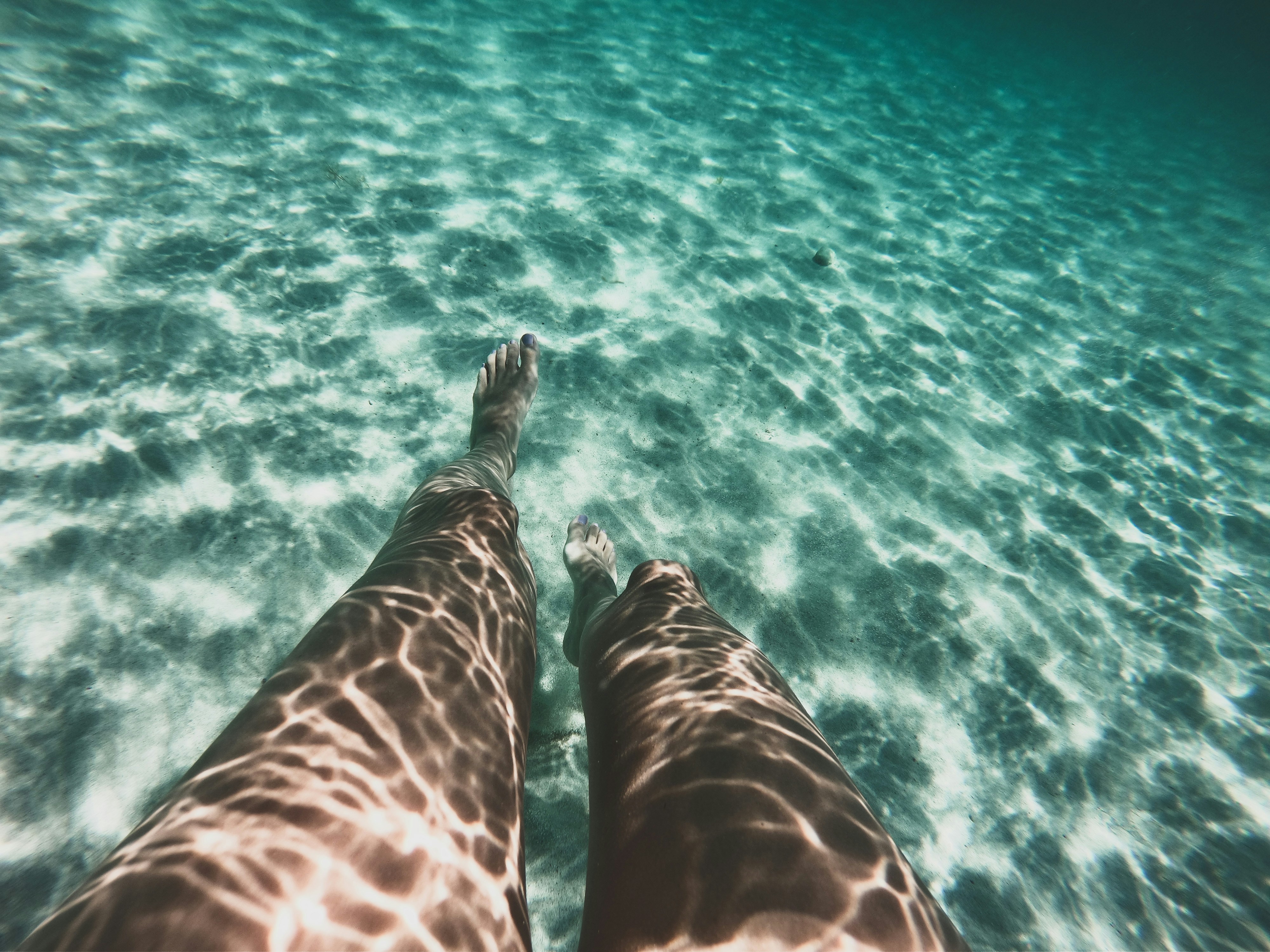 Feet submerged in clear turquoise water, with sunlight creating shimmering patterns on the sandy bottom.