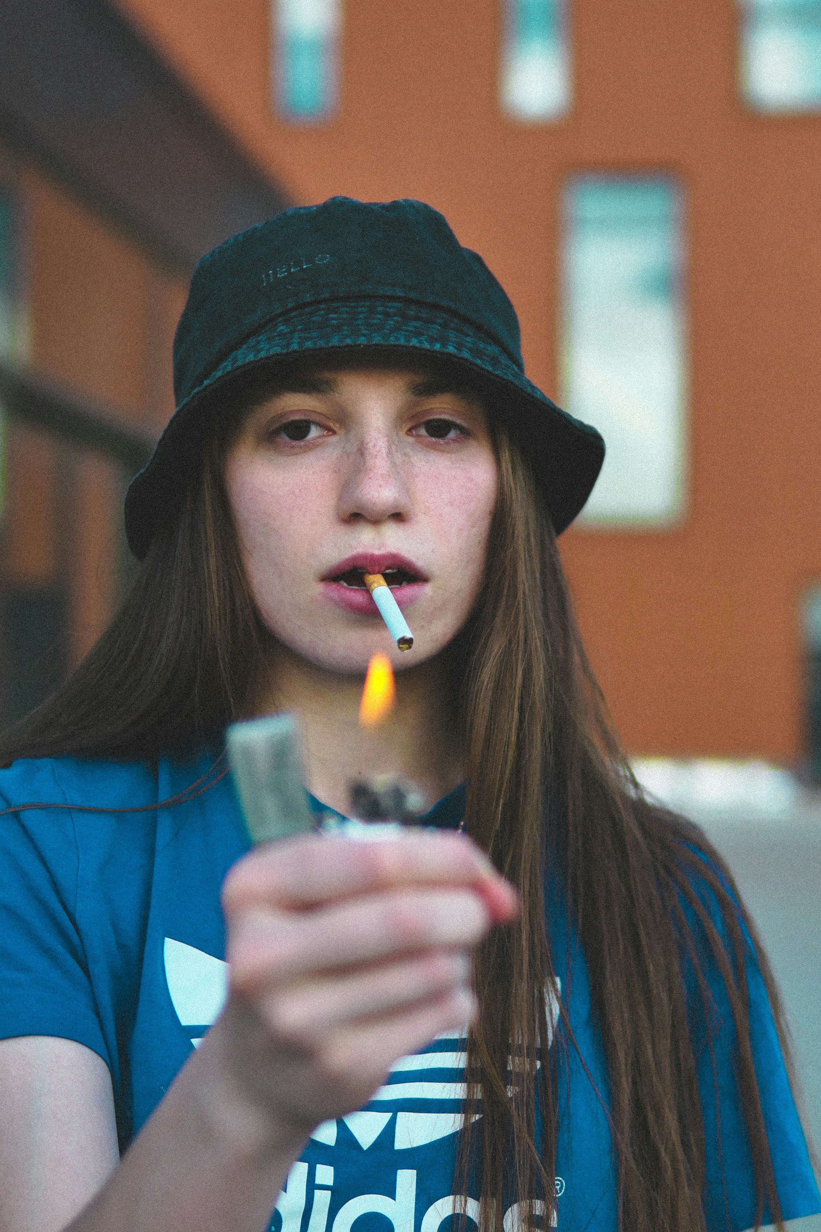Young woman in a blue shirt and hat holding a lit cigarette with a lighter in an urban setting.