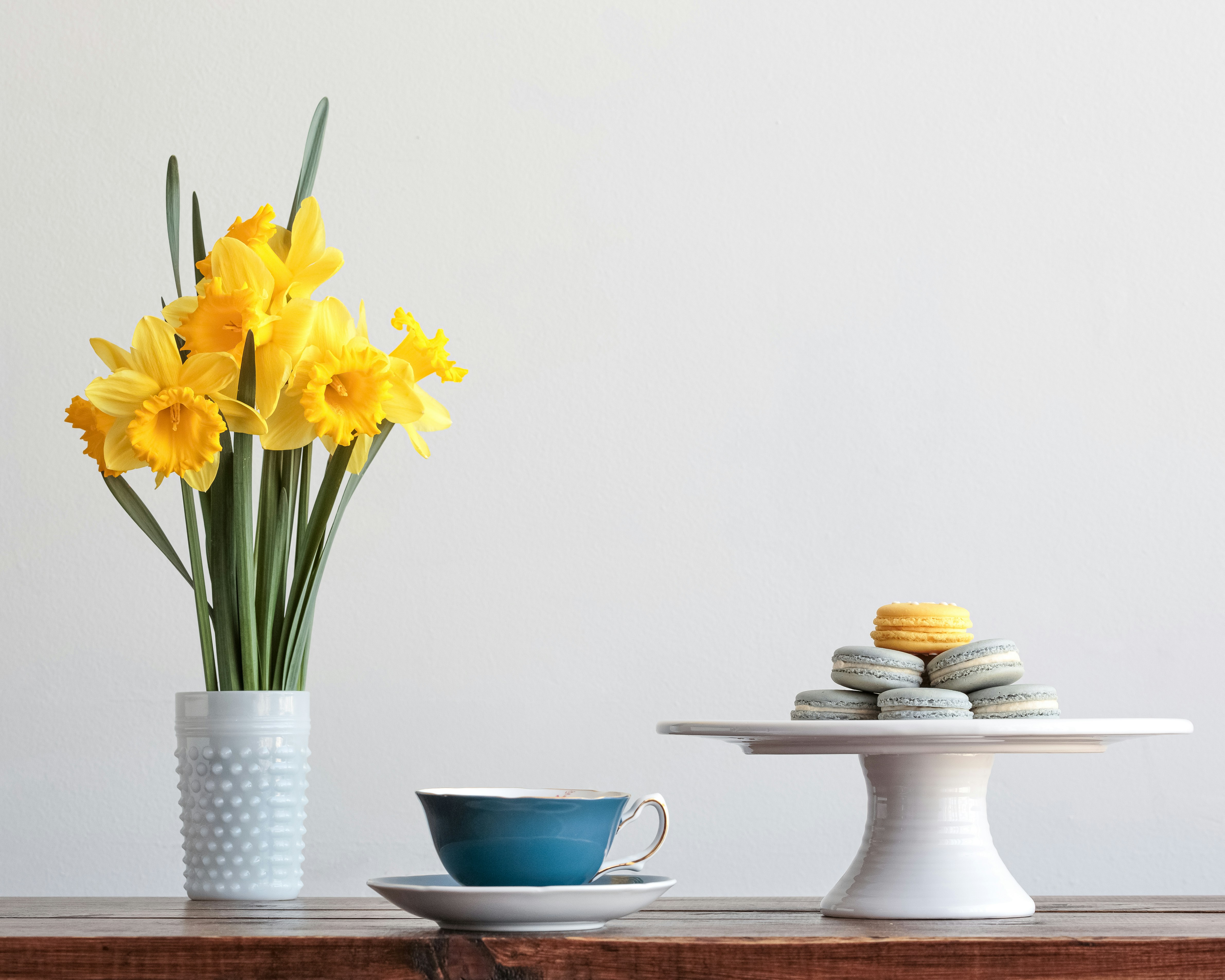 Vibrant daffodils in a textured vase accompany a delicate blue teacup and a tiered stand of pastel macarons, creating a charming table setting.