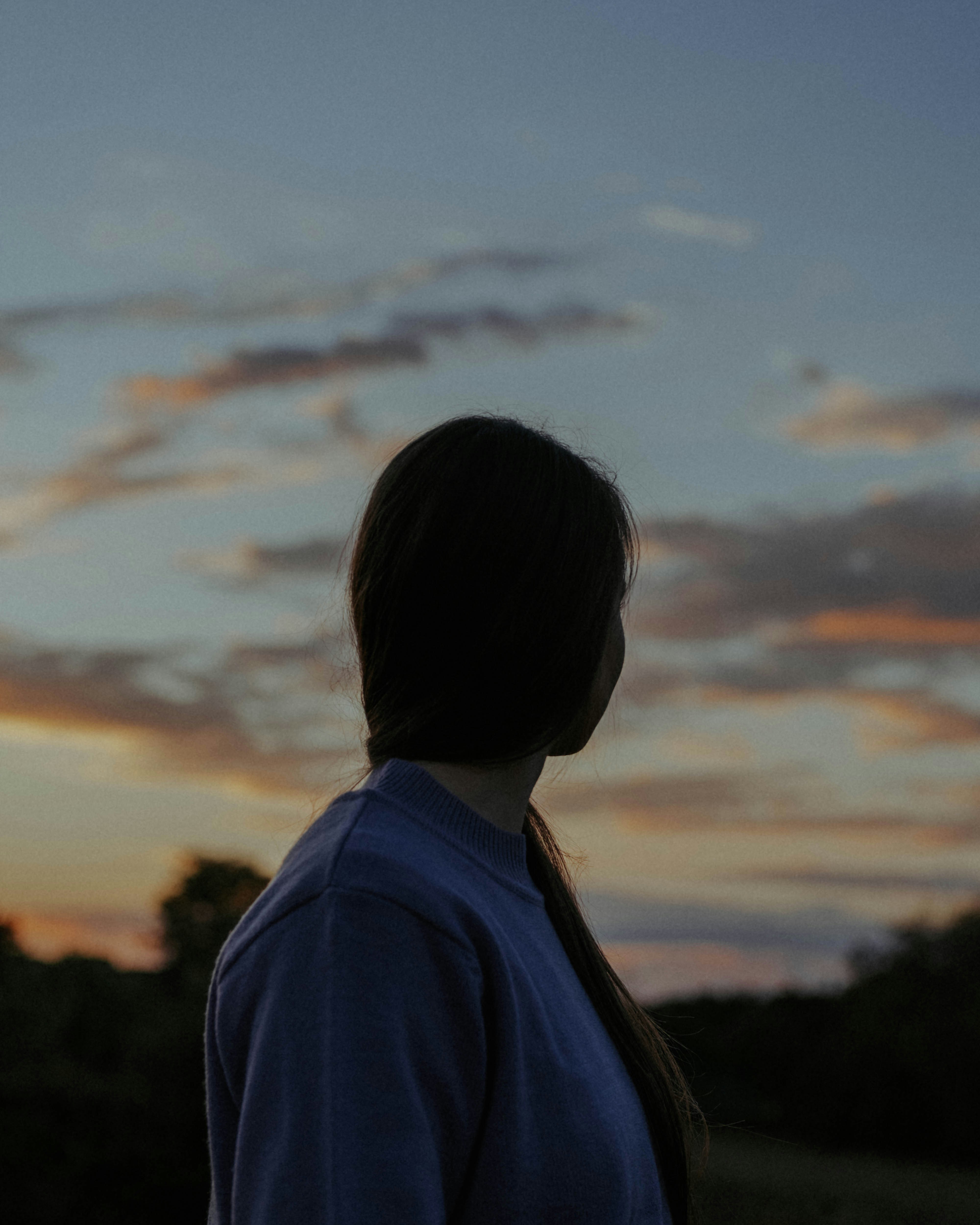 Silhouette of a woman gazing at a colorful sunset, with soft clouds illuminated by the fading light.