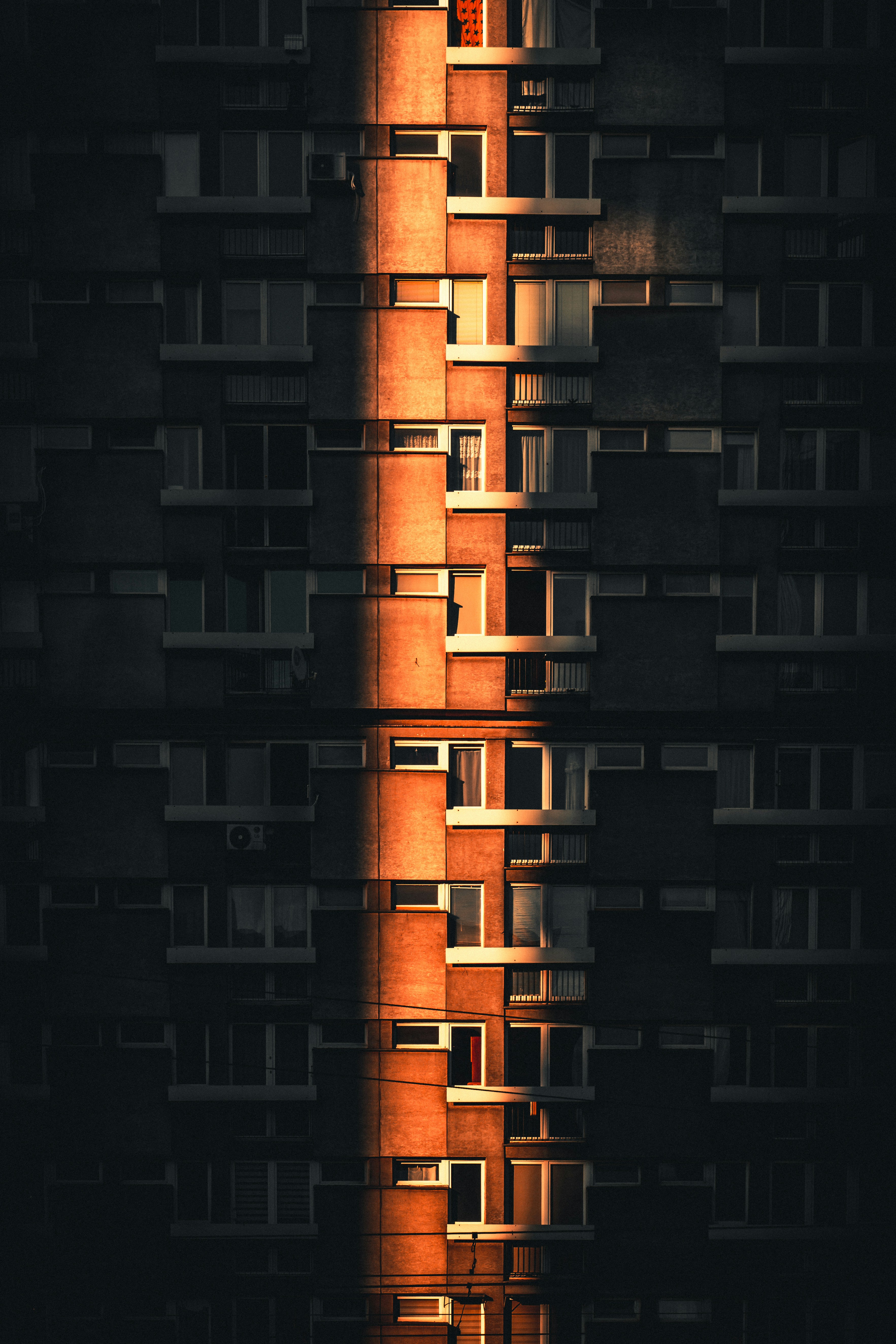 Vertical arrangement of apartment windows illuminated by warm light, contrasting with the dark exterior of the building. The interplay of light creates a striking visual effect.