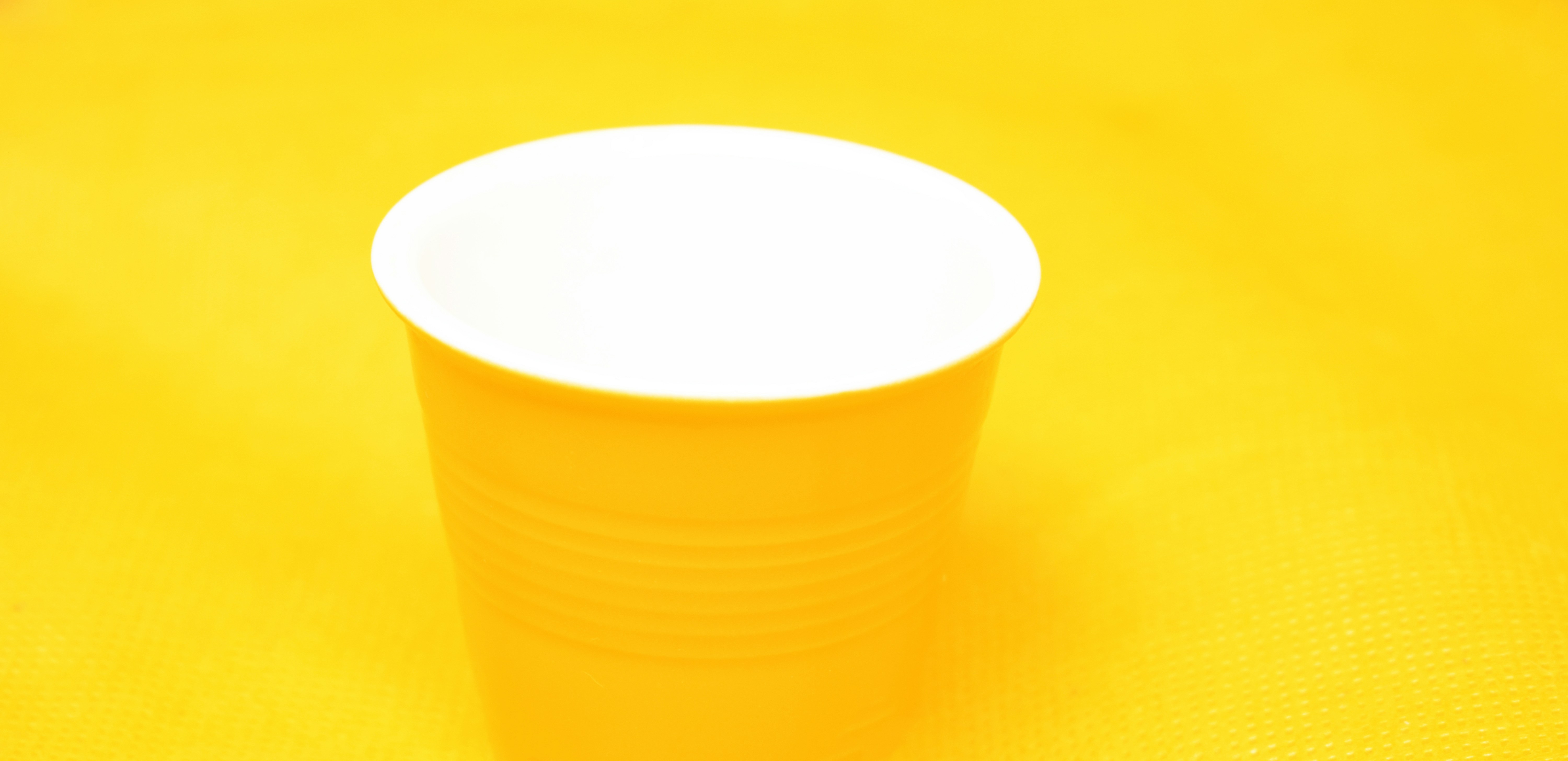 A yellow plastic cup sits against a vibrant yellow background, emphasizing its shape and color. The cup's interior is white, creating a striking contrast.