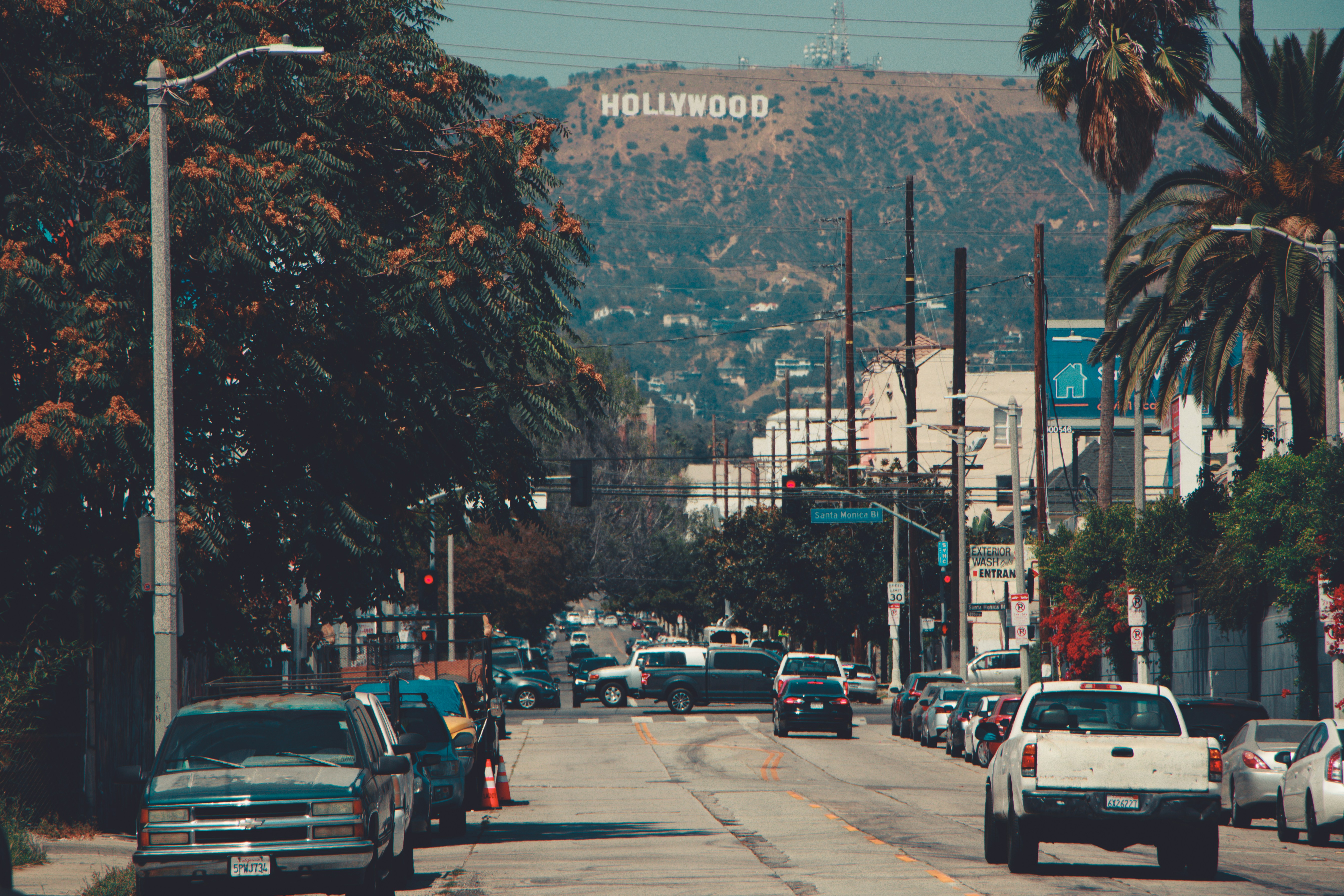 Cars lined along a city street with the Hollywood sign visible on a distant hillside.