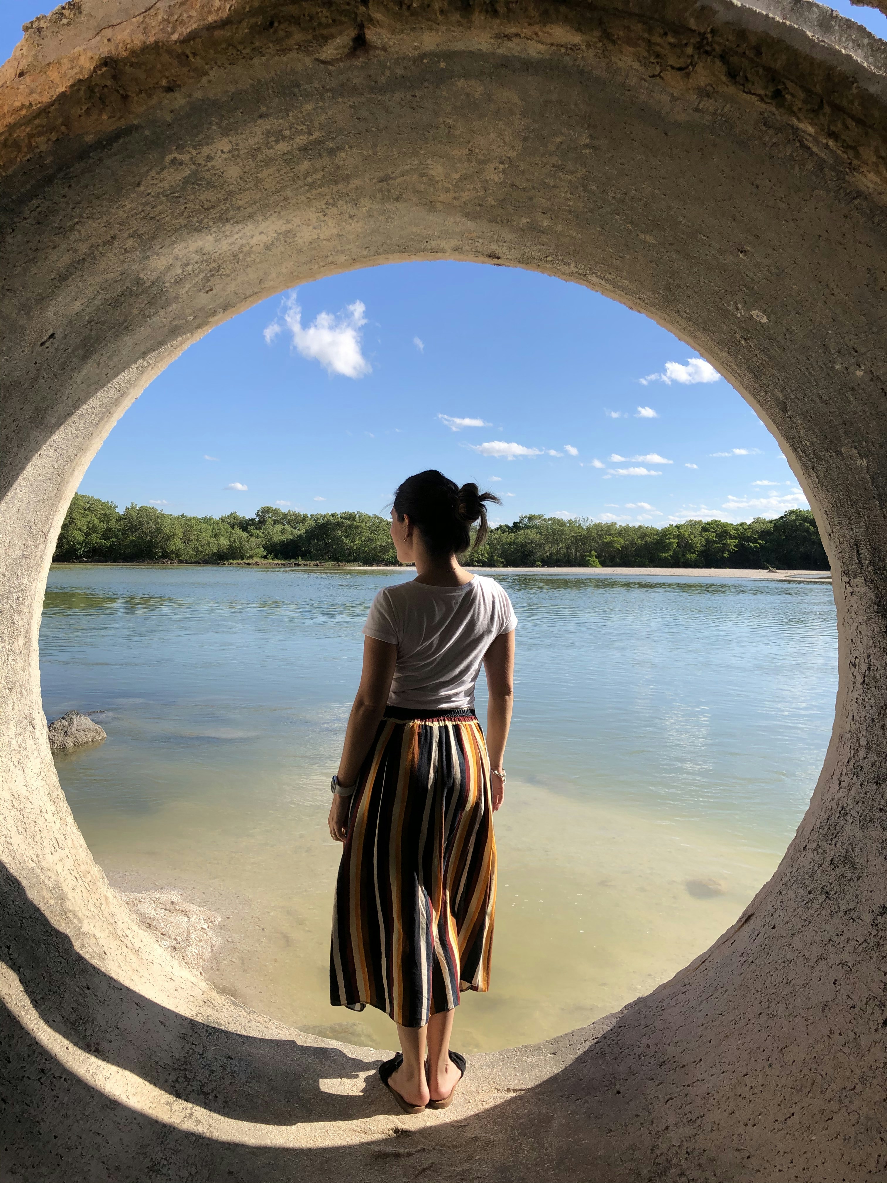 A woman stands in a circular stone frame, gazing at a tranquil river scene under a clear blue sky. The vibrant colors of her striped skirt contrast against the natural backdrop.