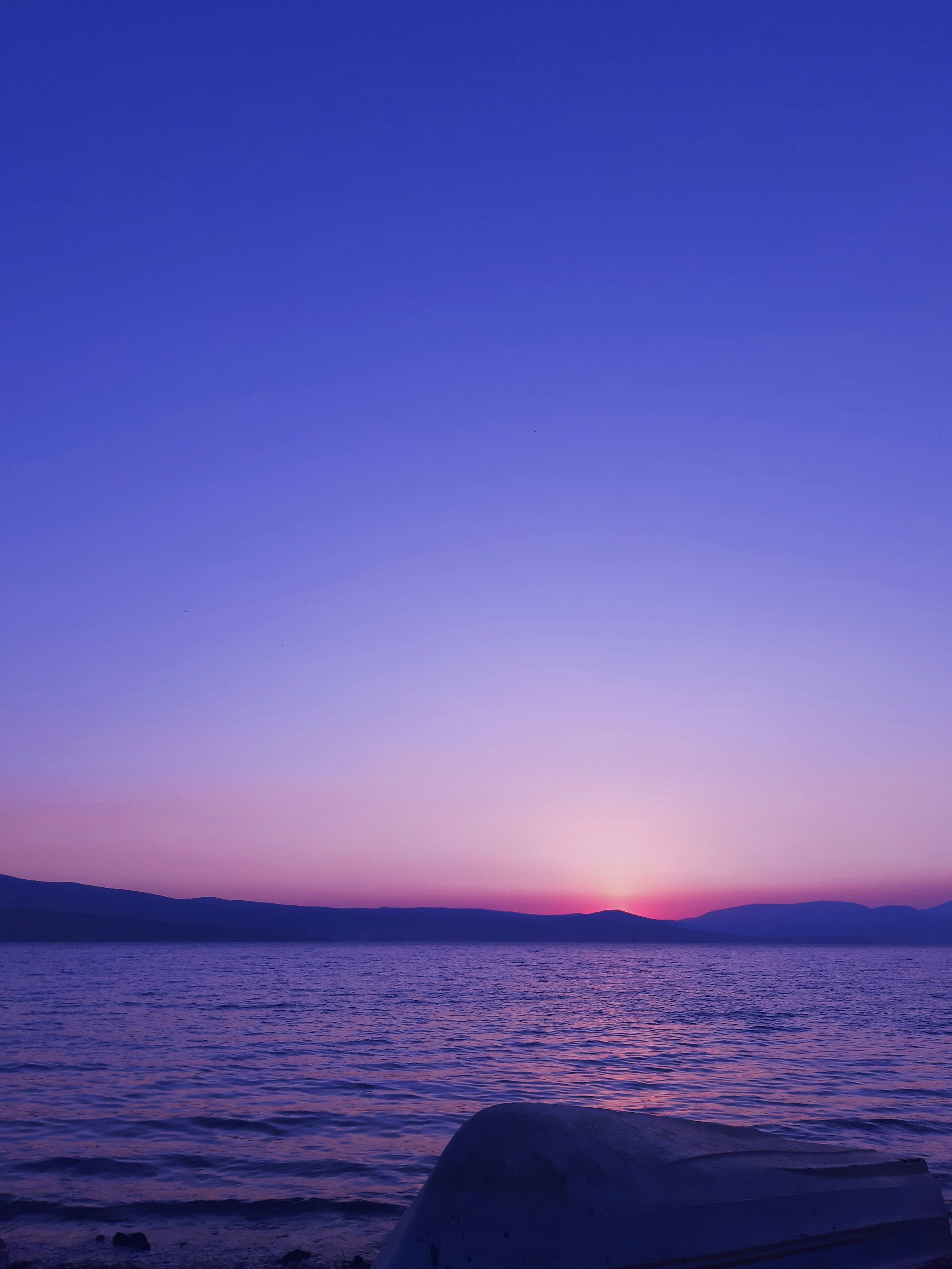 Vibrant gradient of twilight hues reflected on calm water, with distant hills framing the horizon. A peaceful scene capturing the transition from day to night.