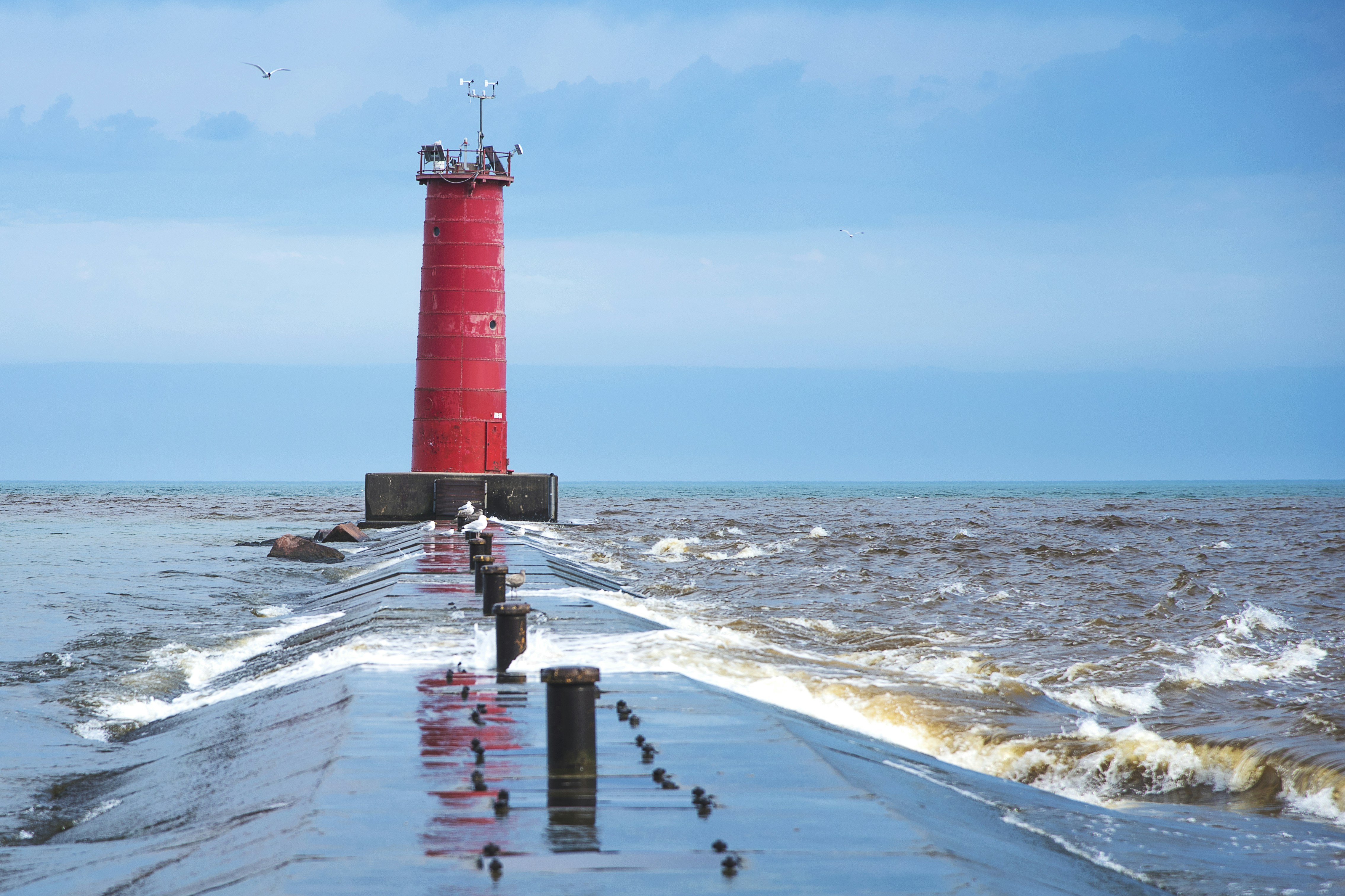 Red lighthouse stands at the end of a pier with waves crashing over the walkway under a bright blue sky.
