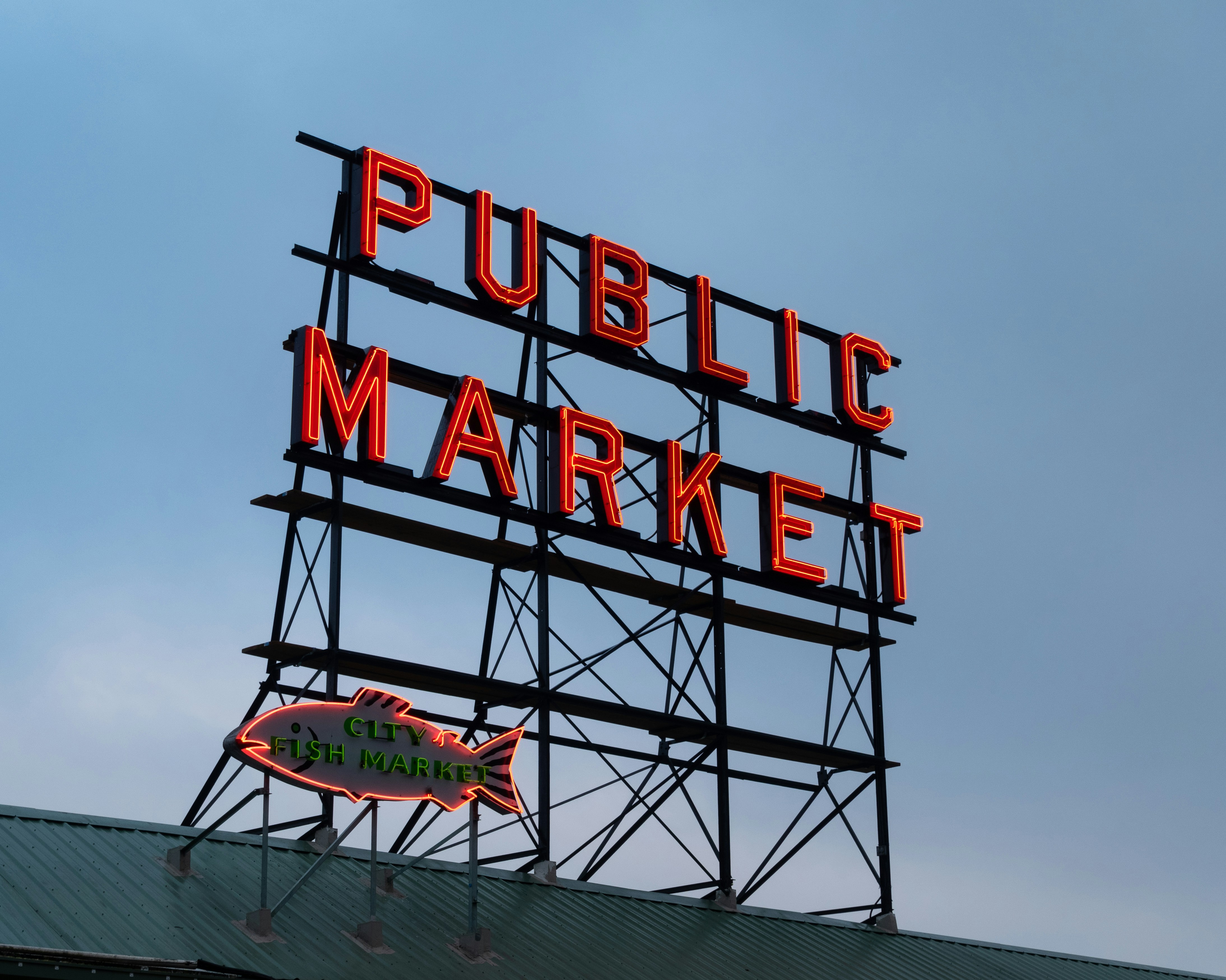 Illuminated sign for a public market featuring bold letters and a fish logo, set against a cloudy sky.