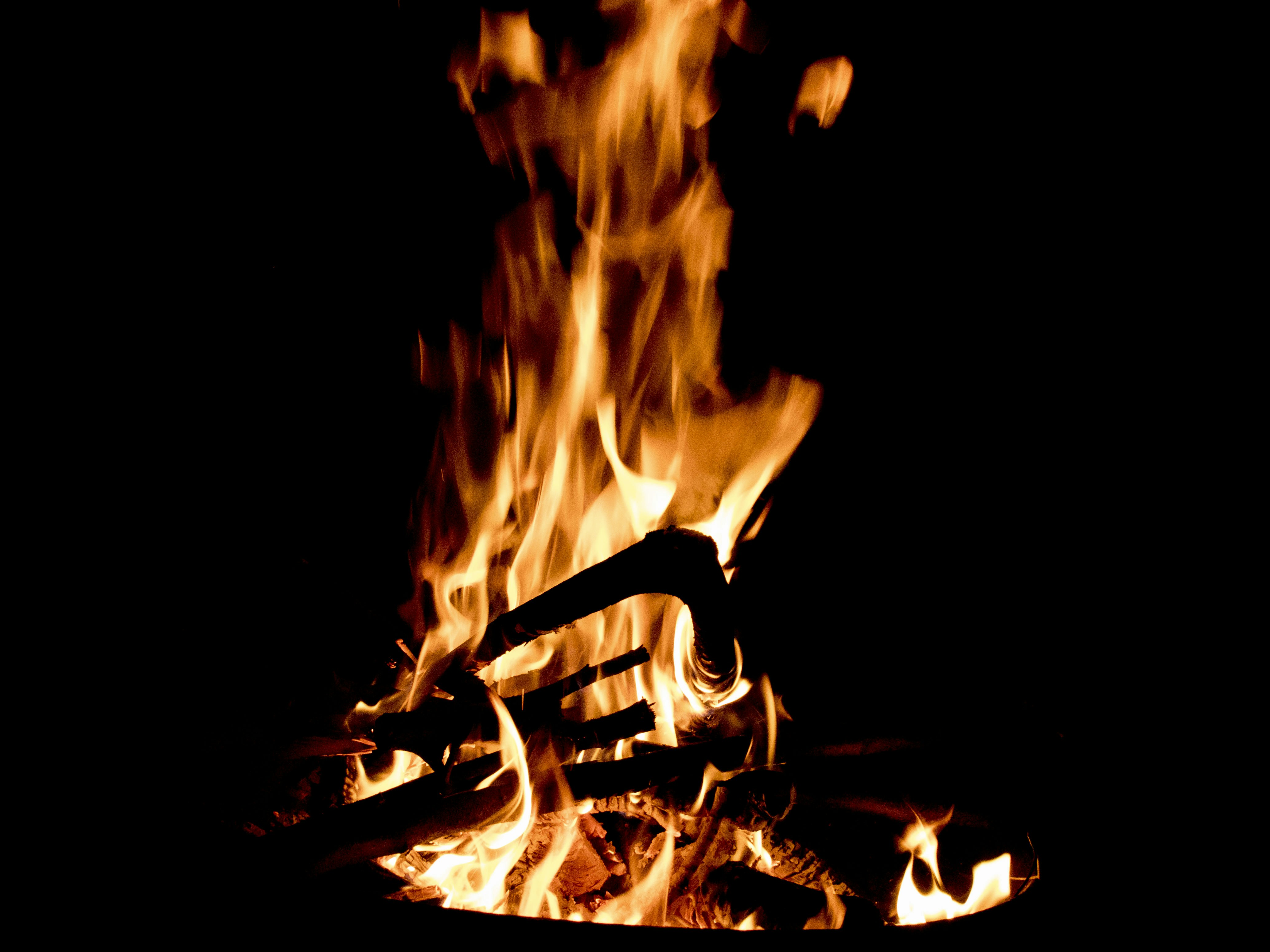 Flickering flames rise from a bed of burning wood, illuminating the darkness with a golden glow.