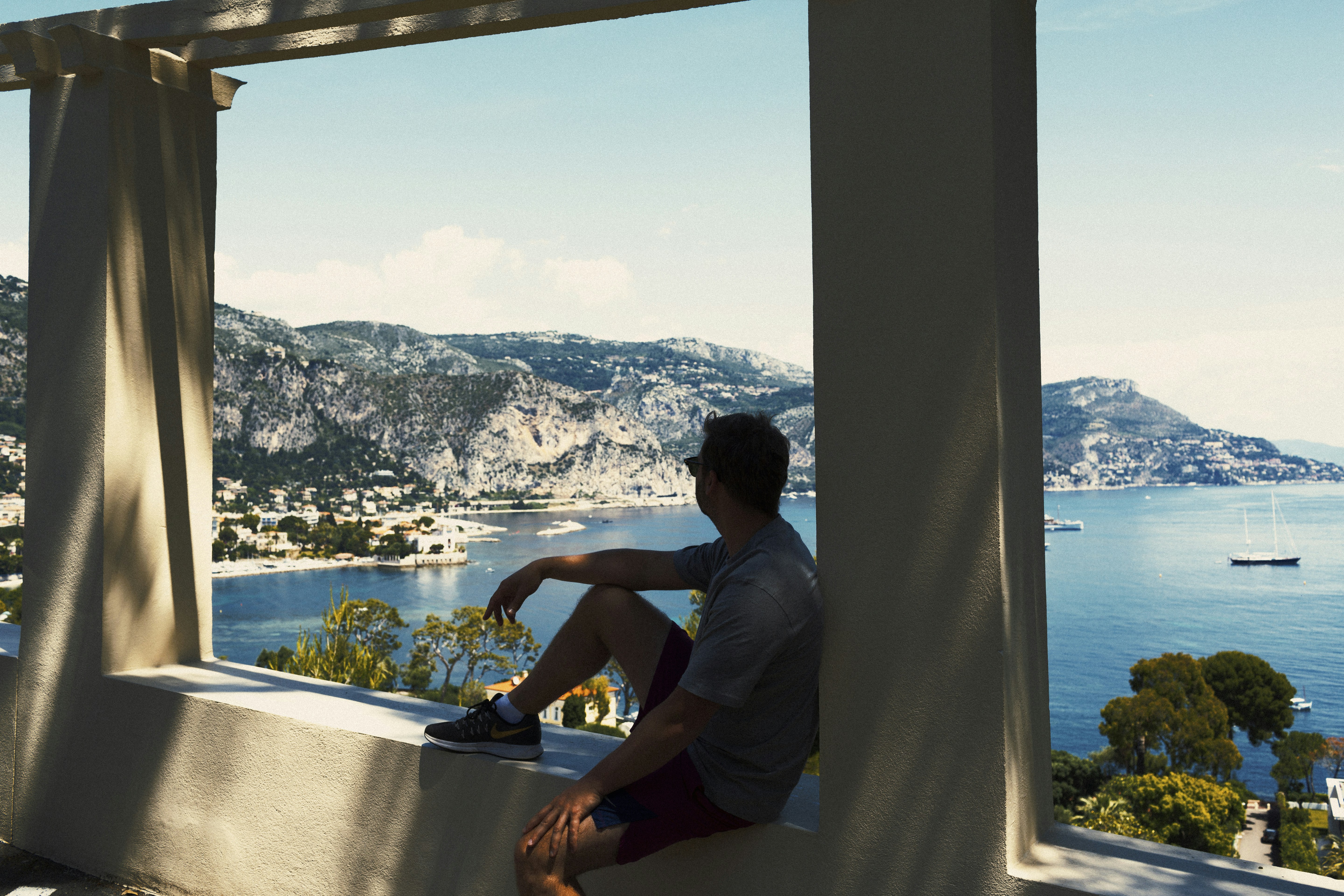 A contemplative figure sits on a ledge, gazing at the serene coastal landscape framed by architectural elements. The scene captures a harmonious blend of nature and structure.