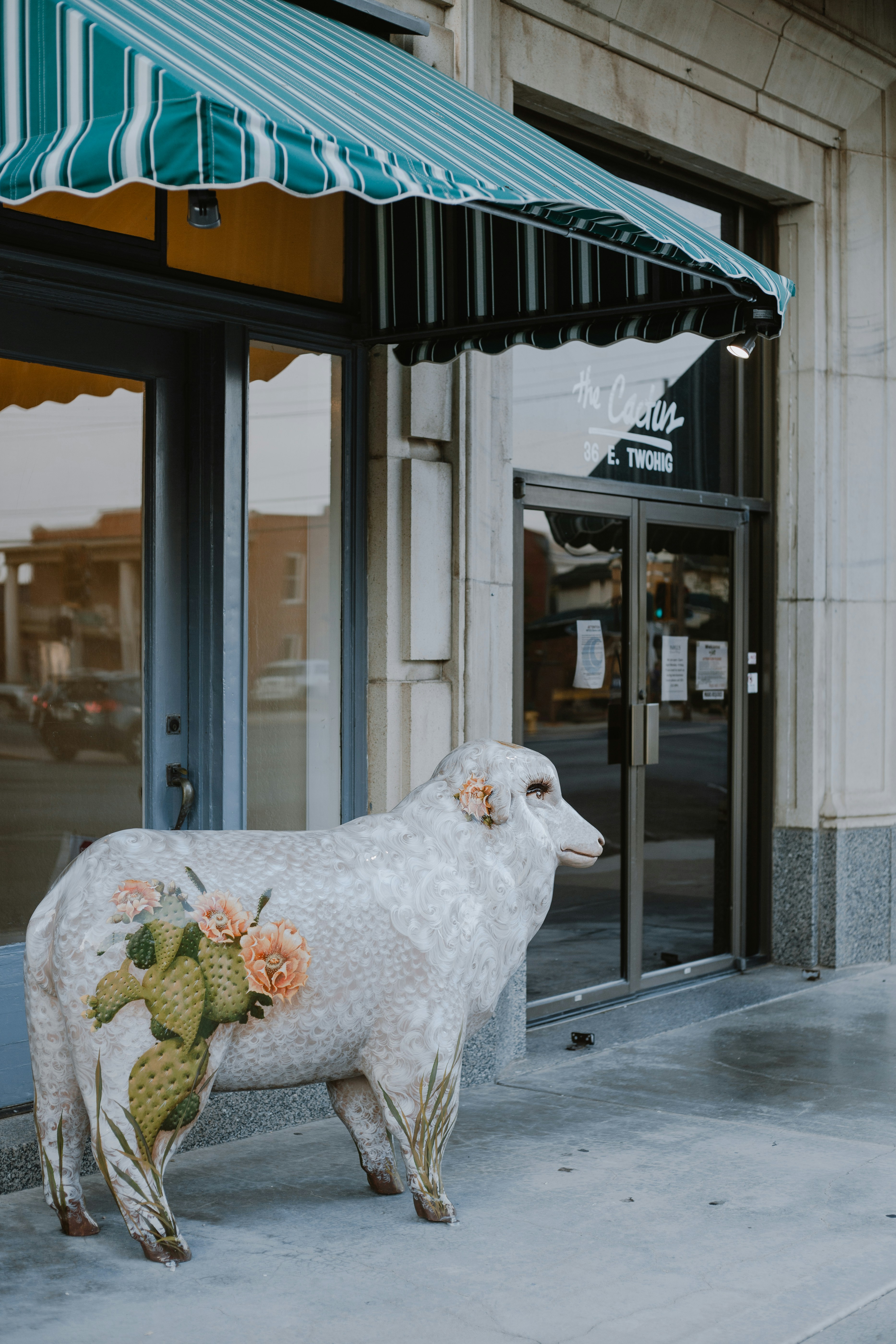 A creatively painted cow sculpture adorned with vibrant flowers and cacti, positioned outside a storefront under a striped awning.