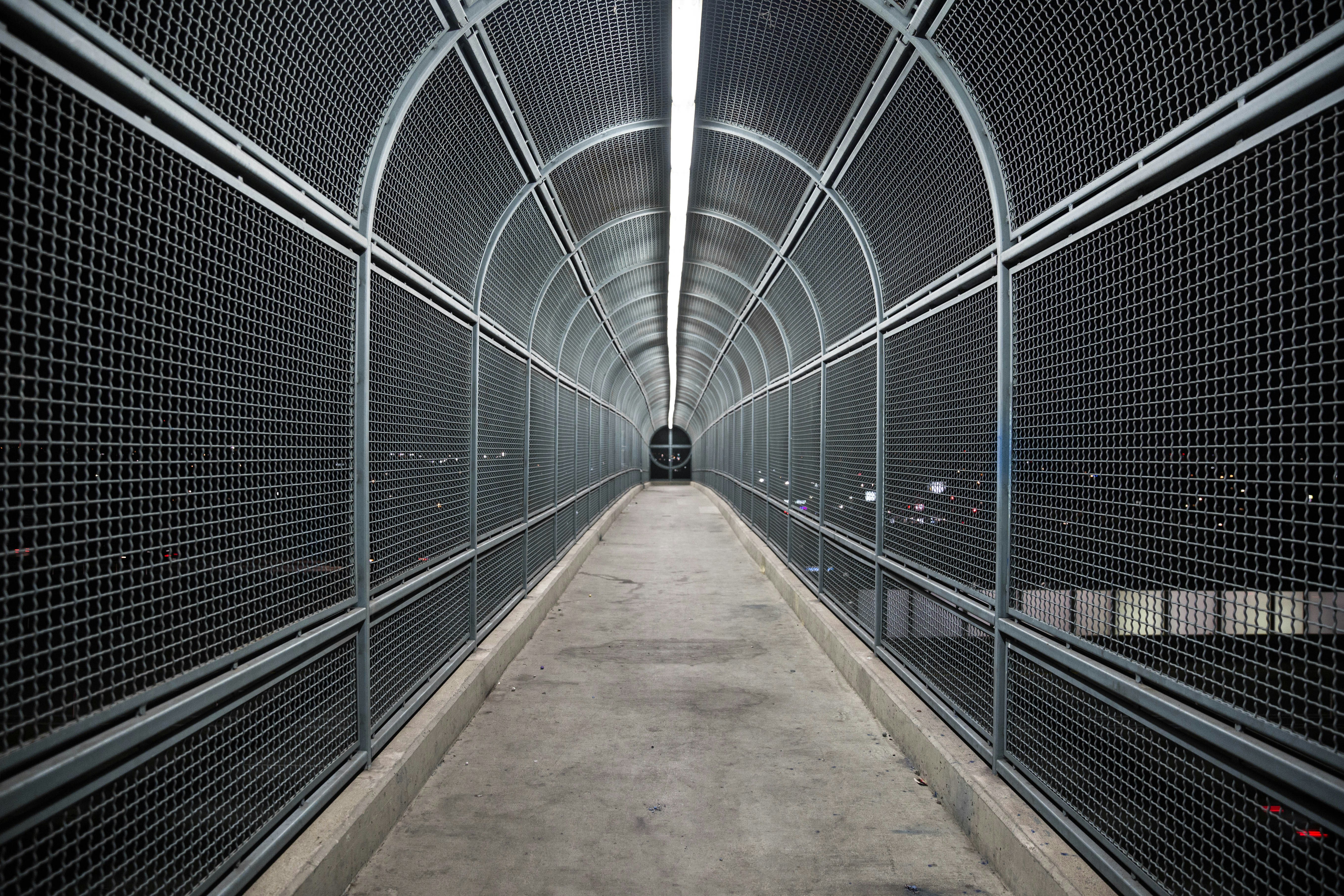 A pedestrian overpass enclosed by a metal mesh structure, leading towards a distant exit illuminated by soft lighting.