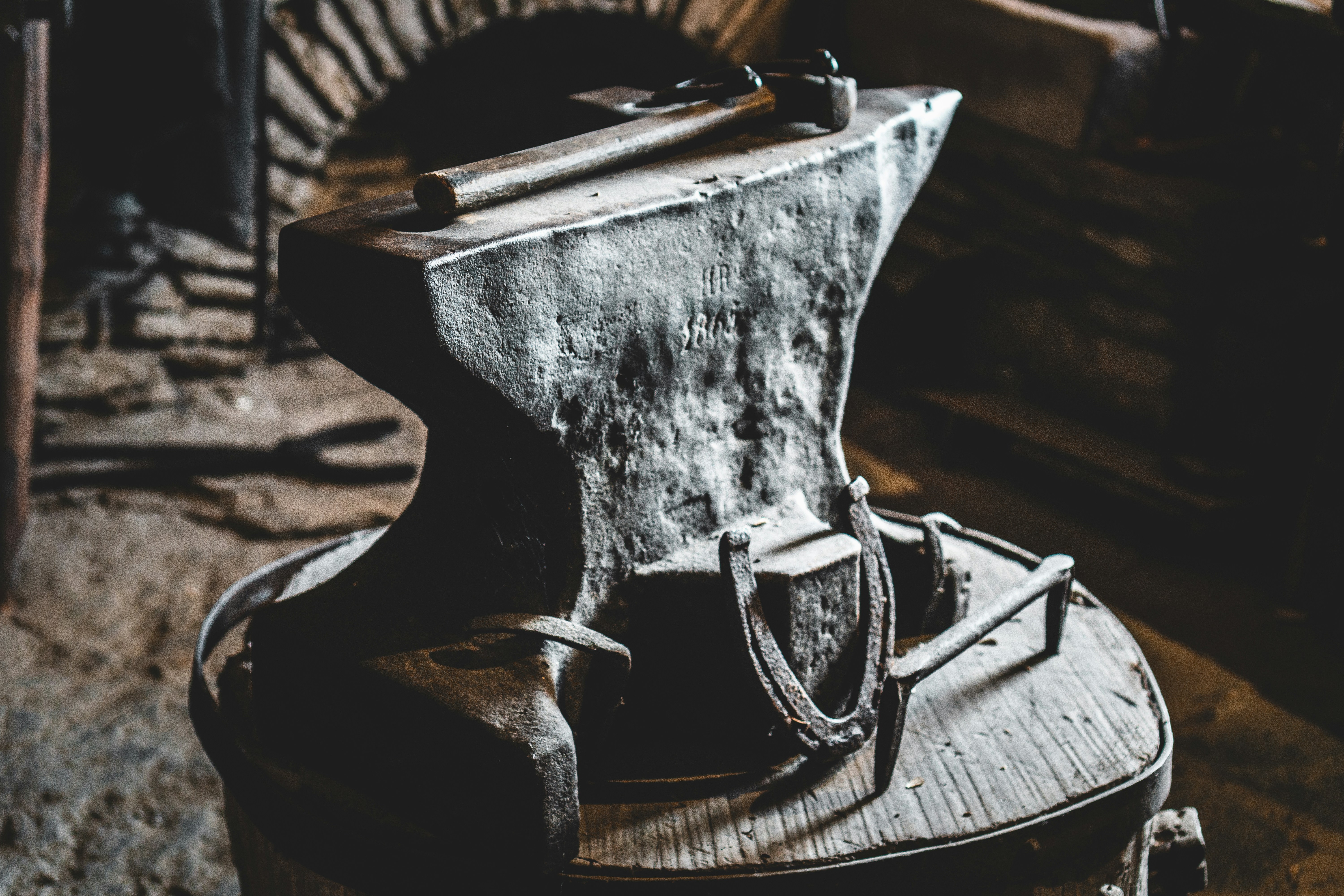 Weathered anvil resting on a wooden surface in a rustic blacksmith shop, surrounded by tools and a stone wall backdrop.