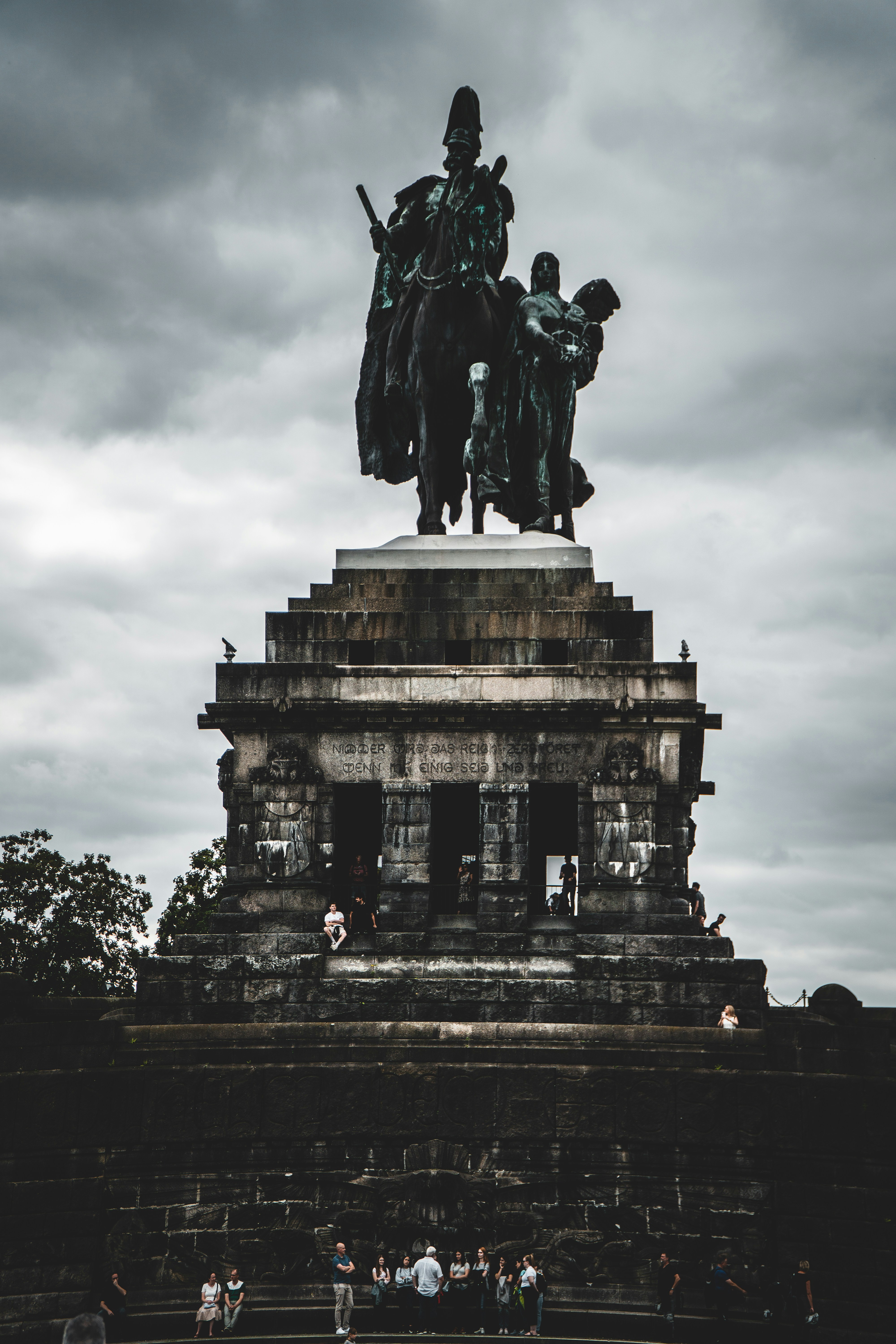 Bronze statue of a mounted figure surrounded by historical figures, set against a moody sky. Visitors gather at the base, emphasizing the monument's grandeur.
