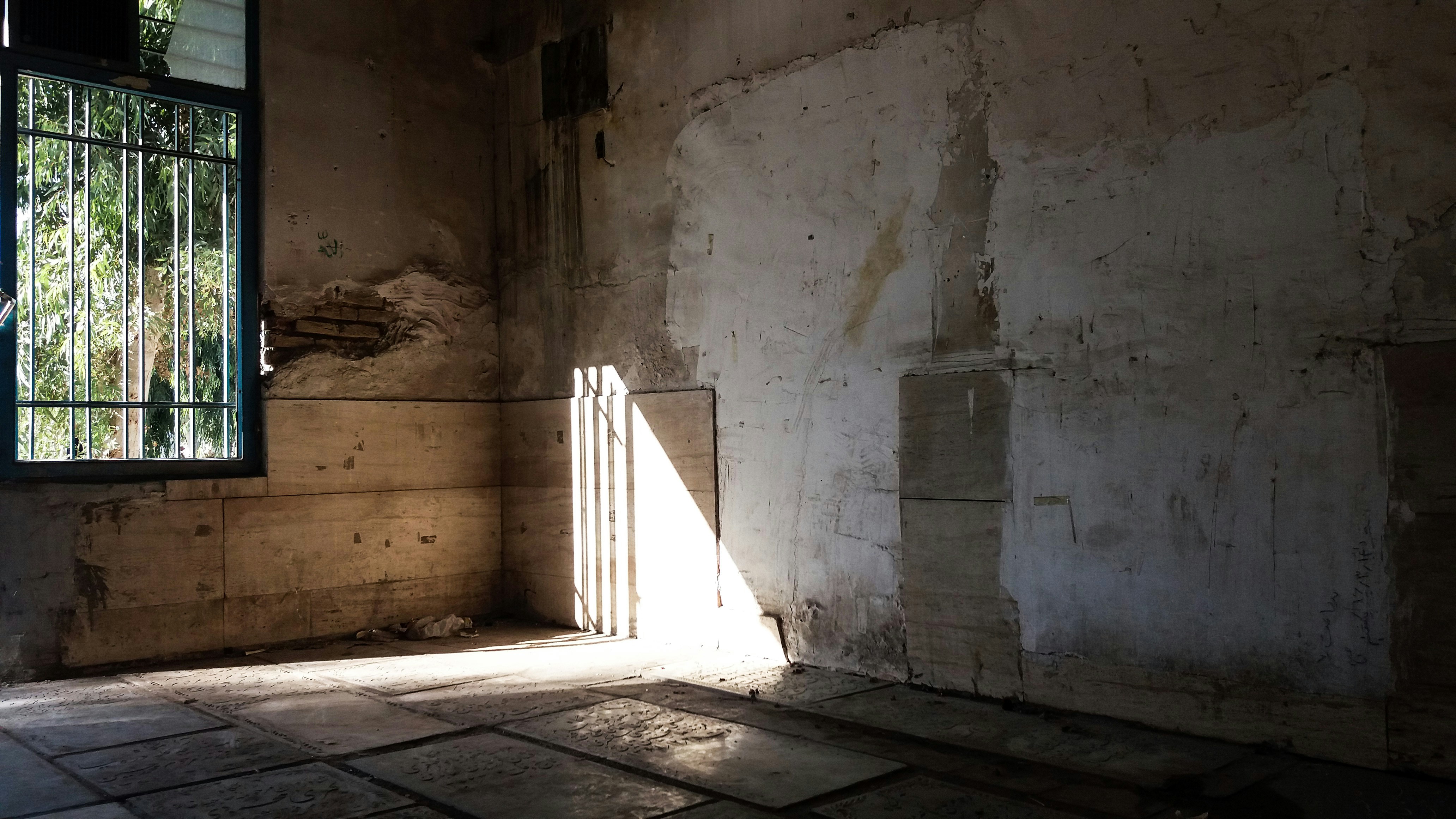 Sunlight streams through a barred window, illuminating the dusty floor of an empty room with peeling walls. The interplay of light and shadow evokes a sense of nostalgia.