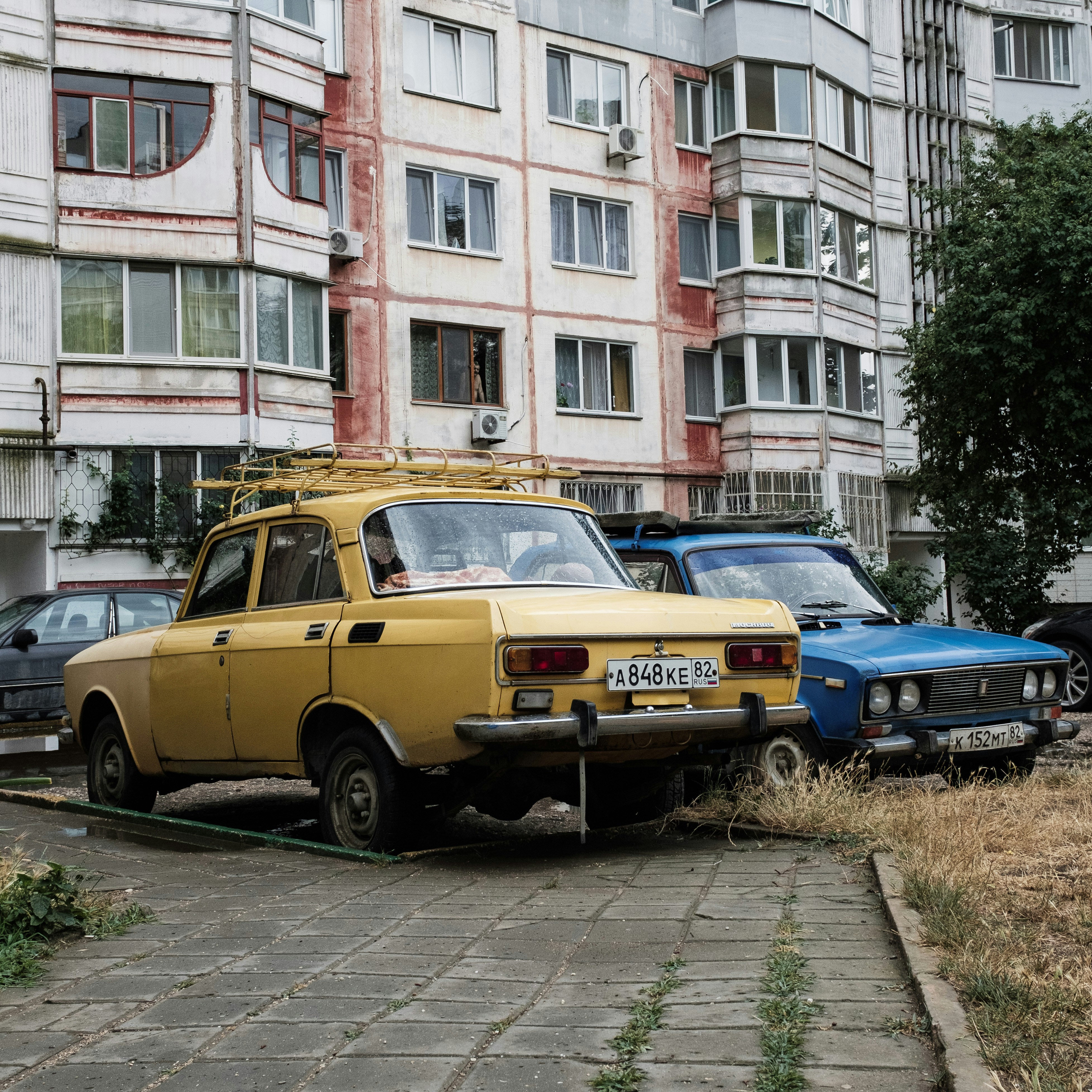 Two vintage cars parked outside a weathered apartment building with distinctive Soviet-era architecture.