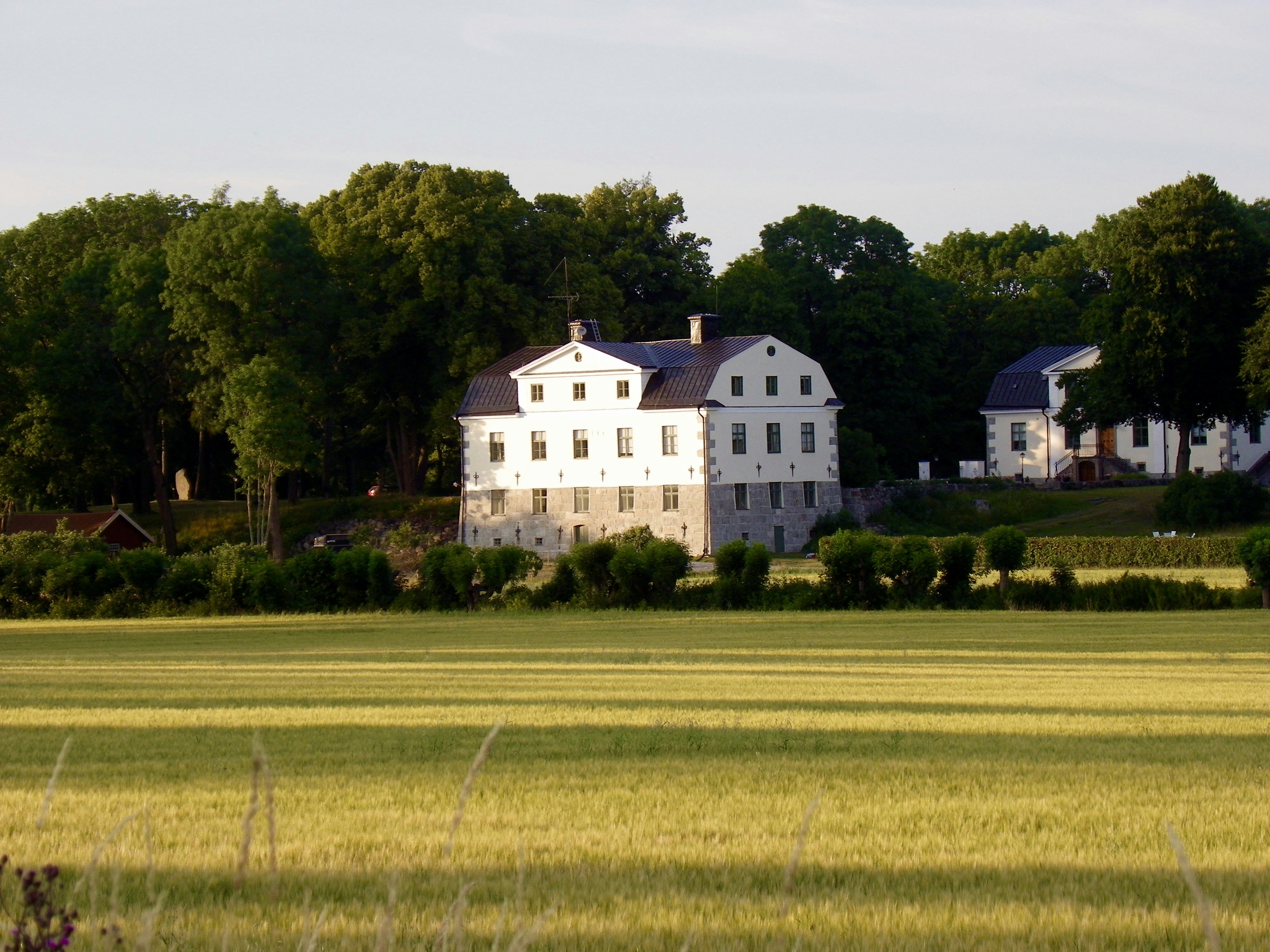A historic white house nestled among verdant trees, overlooking expansive golden fields under a clear sky.